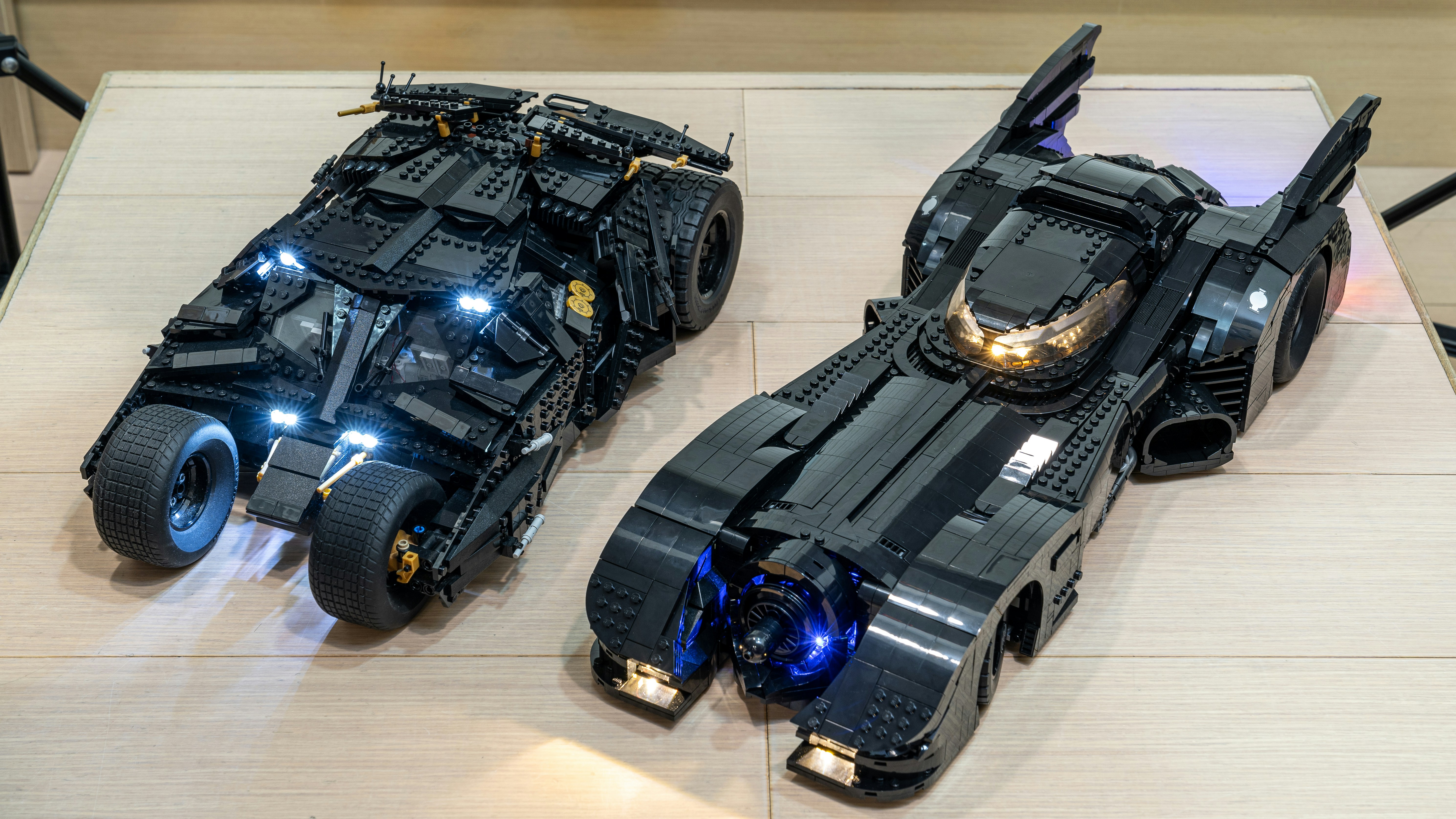 Two detailed model Batmobiles with illuminated headlights on a wooden surface.
