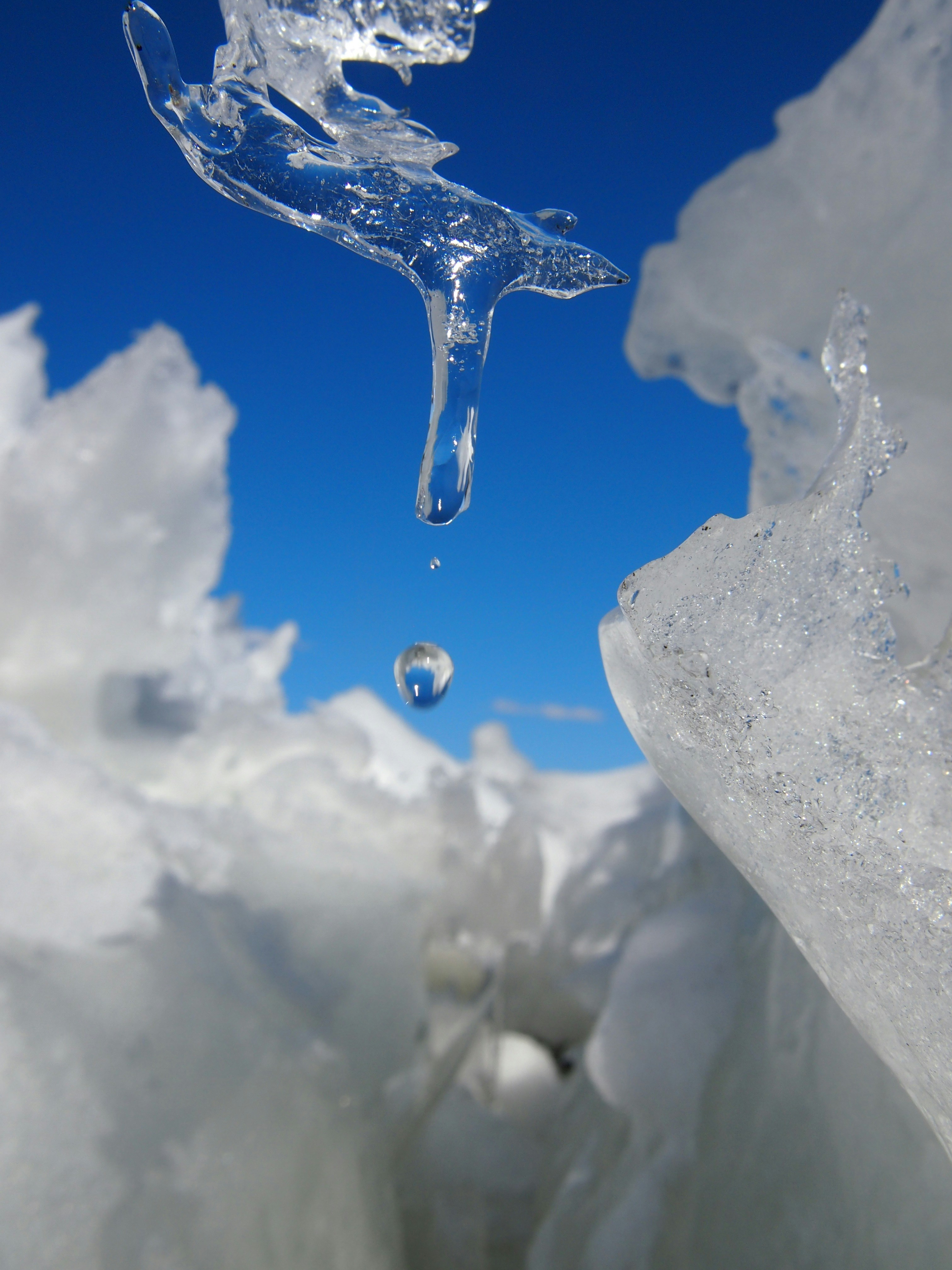 Close-up photograph of a hanging ice formation with a droplet about to fall, set against a bright blue sky and sculpted snow walls.