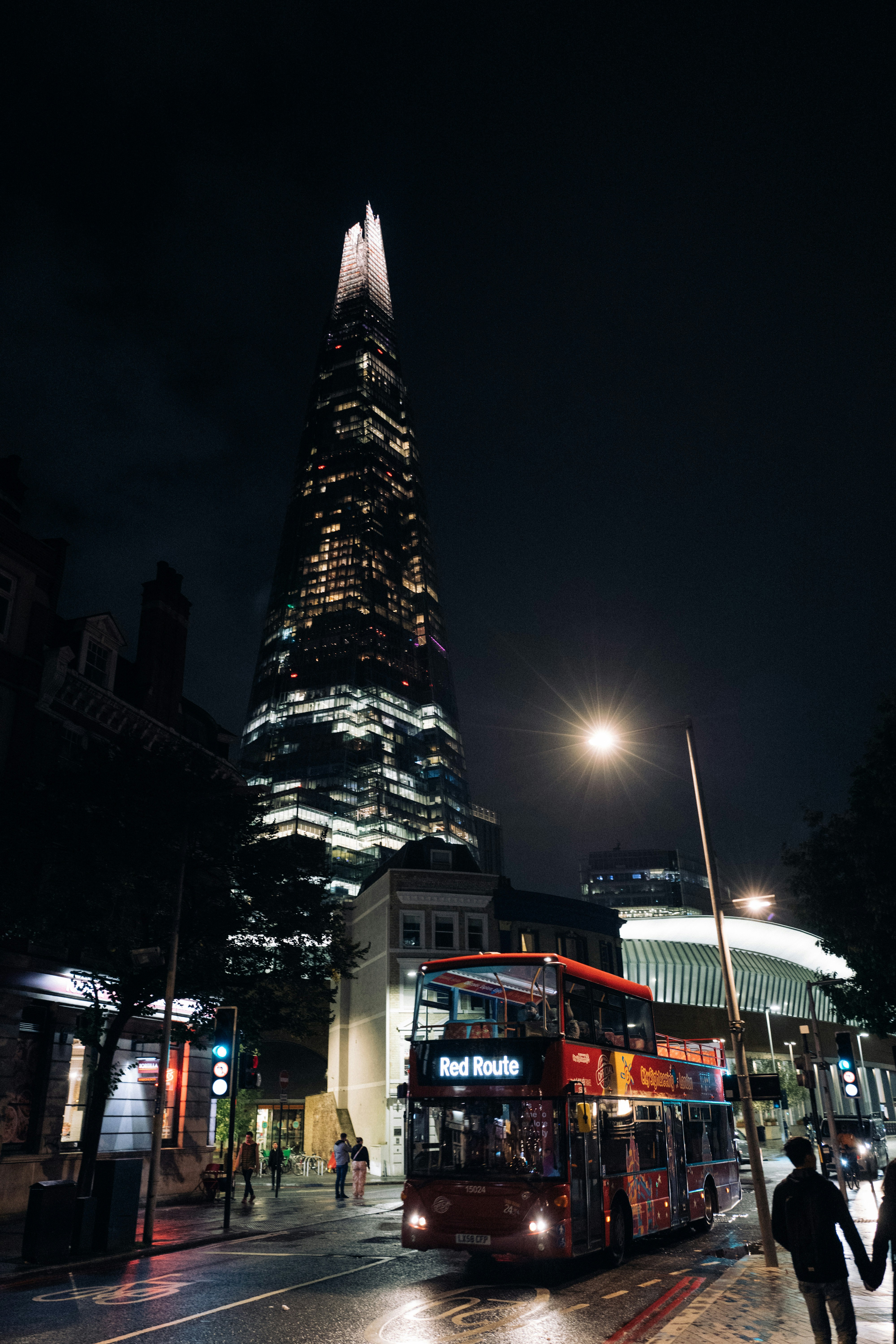 Night photograph of the Shard towering above a red double-decker bus on a lively London street, lit by streetlights. The scene captures urban energy with moody skies and late-night pedestrians.