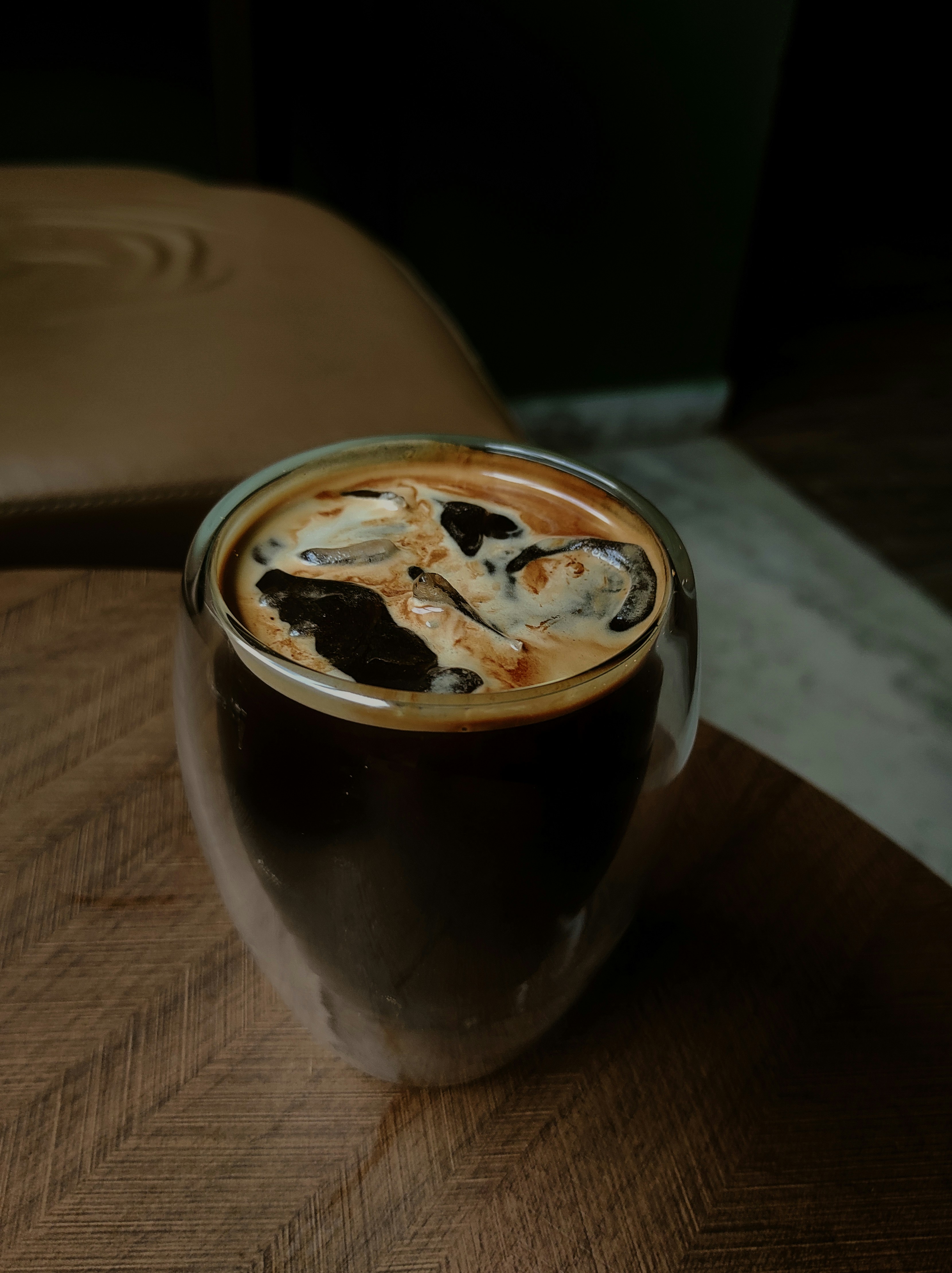 Iced espresso in a curved tulip glass rests on a rustic wooden table, with ice cubes visible in the crema. The dim interior provides a warm, intimate backdrop.