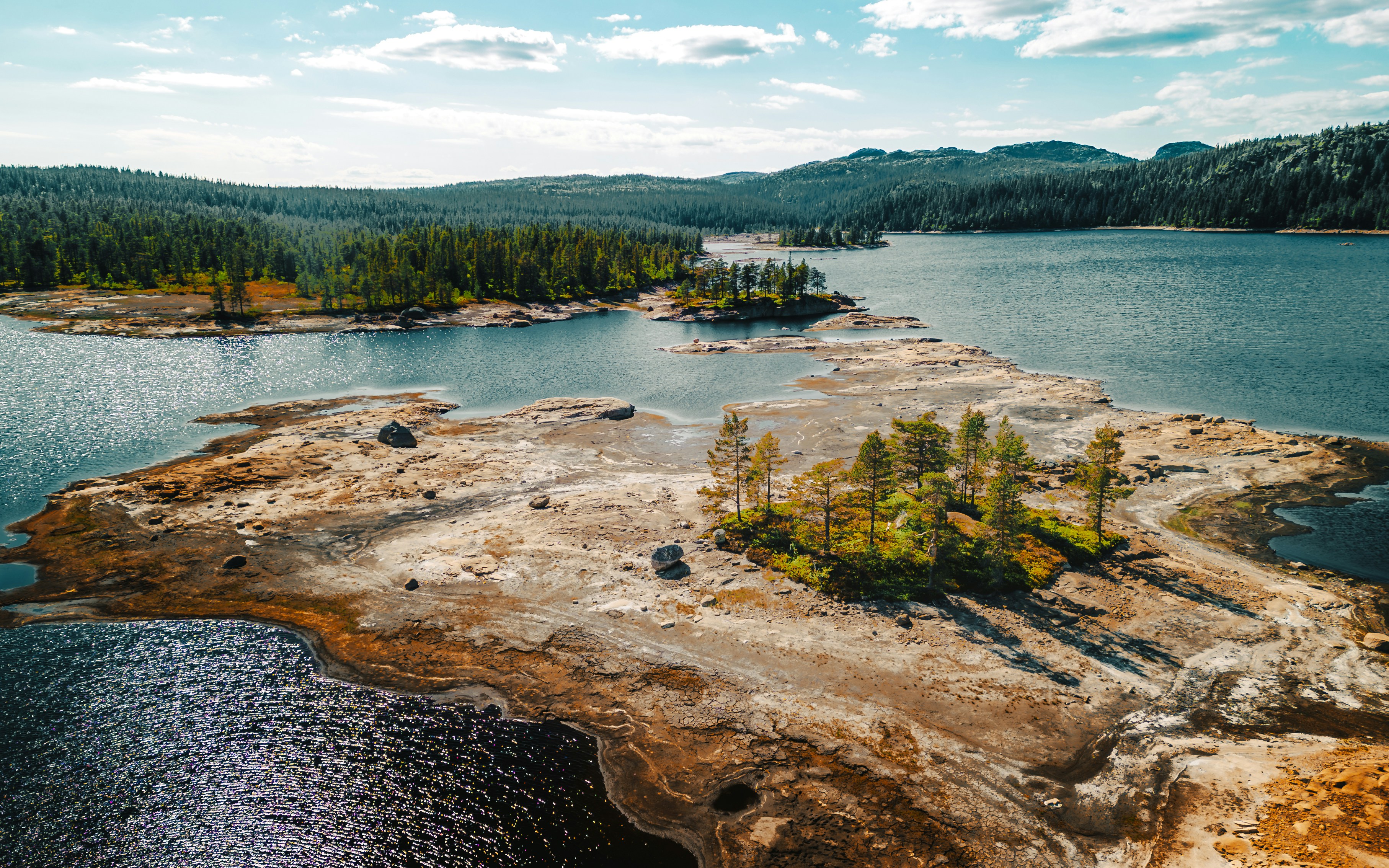 Aerial view of a small forested peninsula surrounded by a tranquil lake and distant mountains under a blue sky.