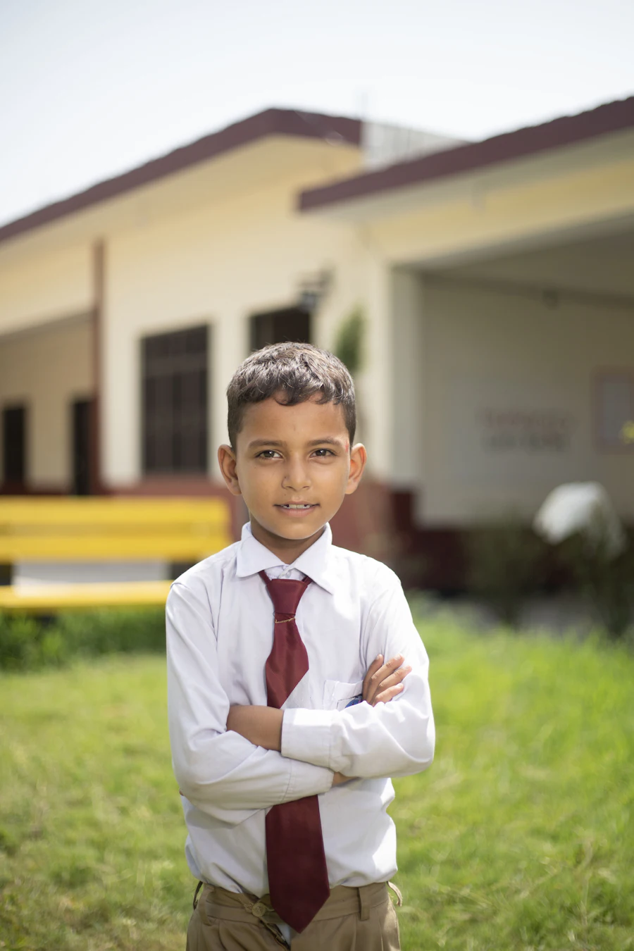 A young student in a white shirt and burgundy tie stands with arms crossed on a sunny schoolyard, the building and greenery softly blurred in the background. (Photo, likely a school portrait or candid exterior shot).