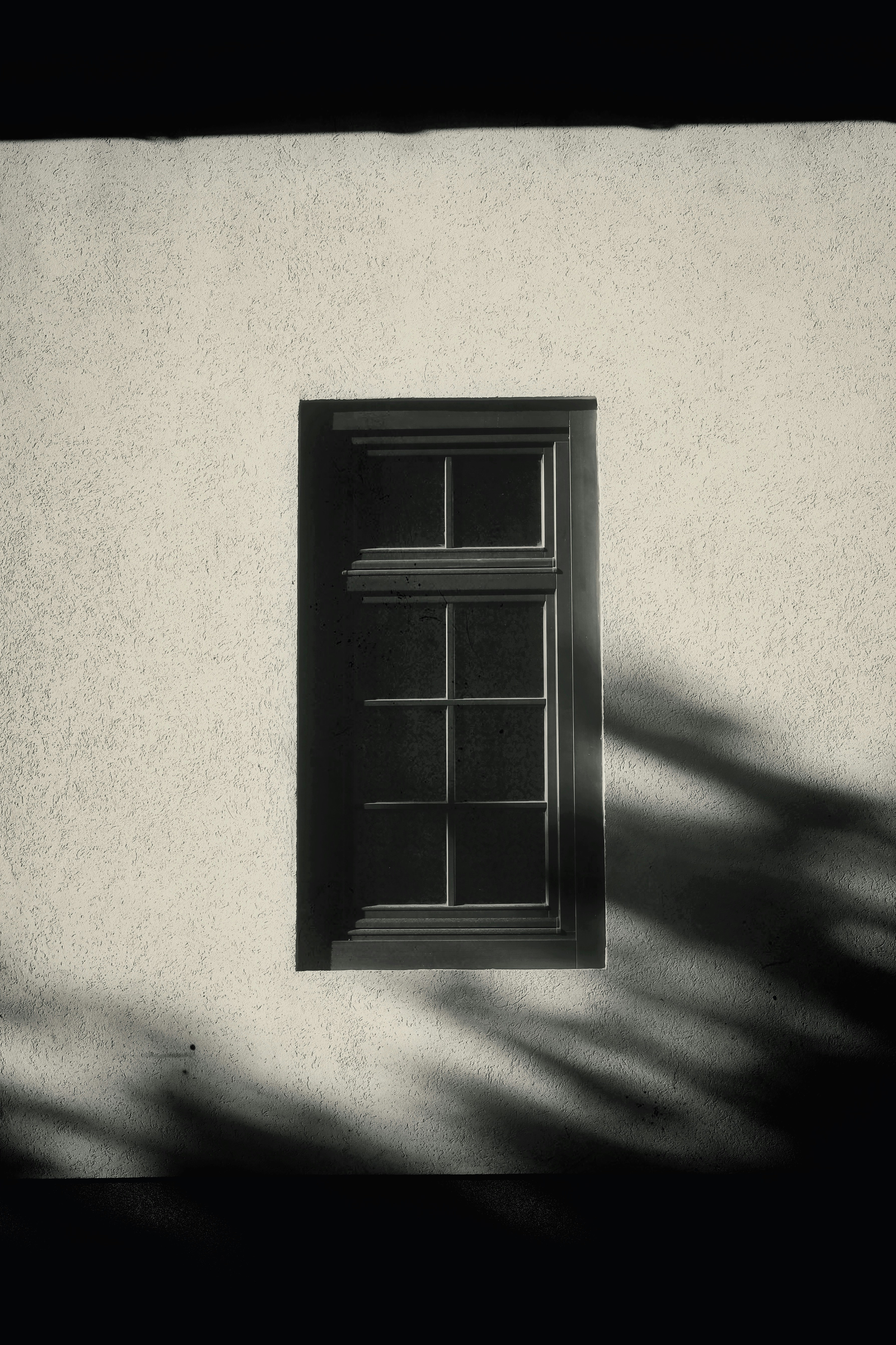 Grayscale exterior wall with a narrow window; bold shadows sweep across the stucco, highlighting texture and geometry.