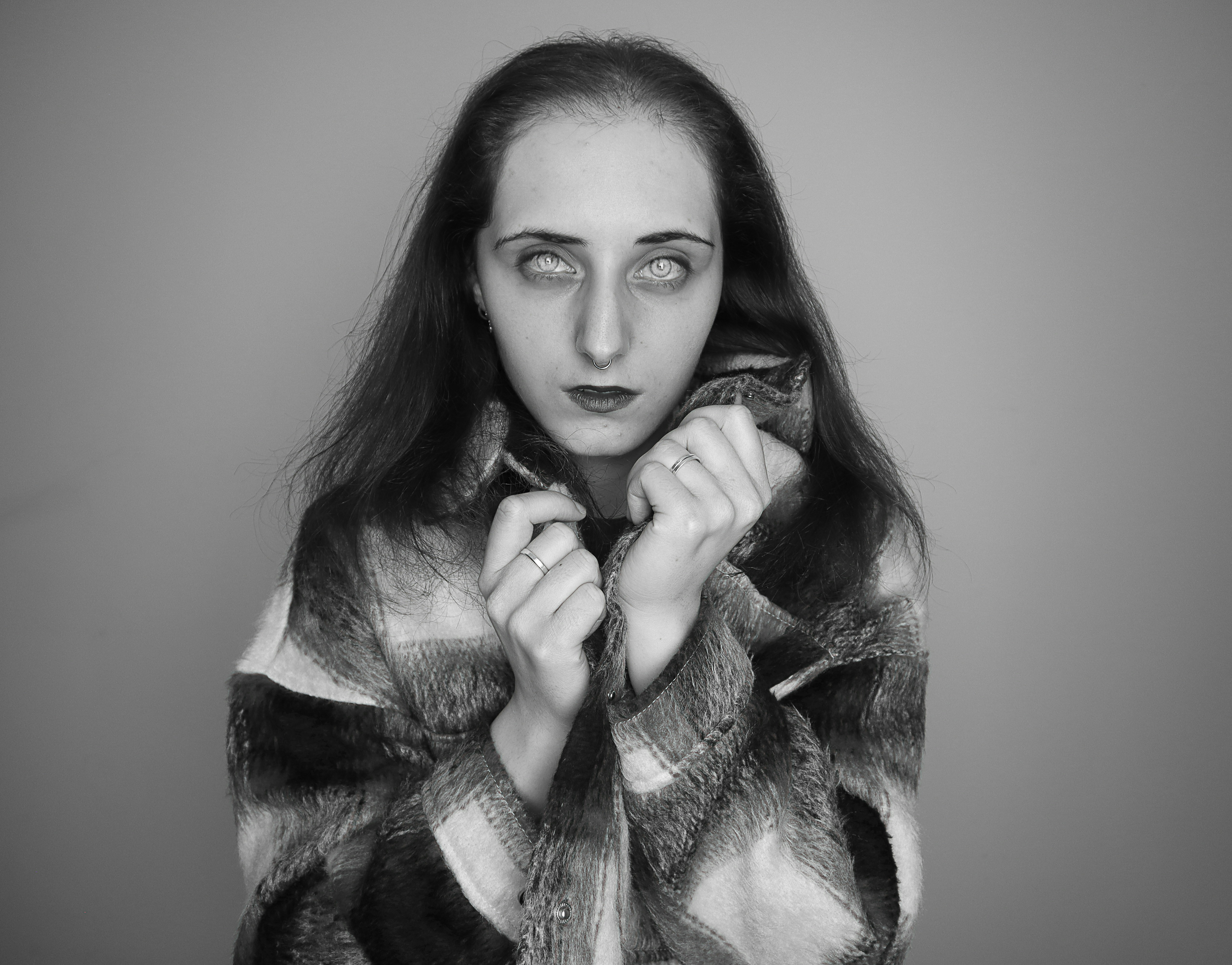 This captivating black and white portrait features a subject with an intense gaze, enveloped in a textured, plaid coat. The composition centers on the subject's eyes, creating a dramatic effect enhanced by the stark contrasts of light and shadow. The grayscale palette and soft lighting evoke a hauntingly serene atmosphere, making the image both striking and memorable.