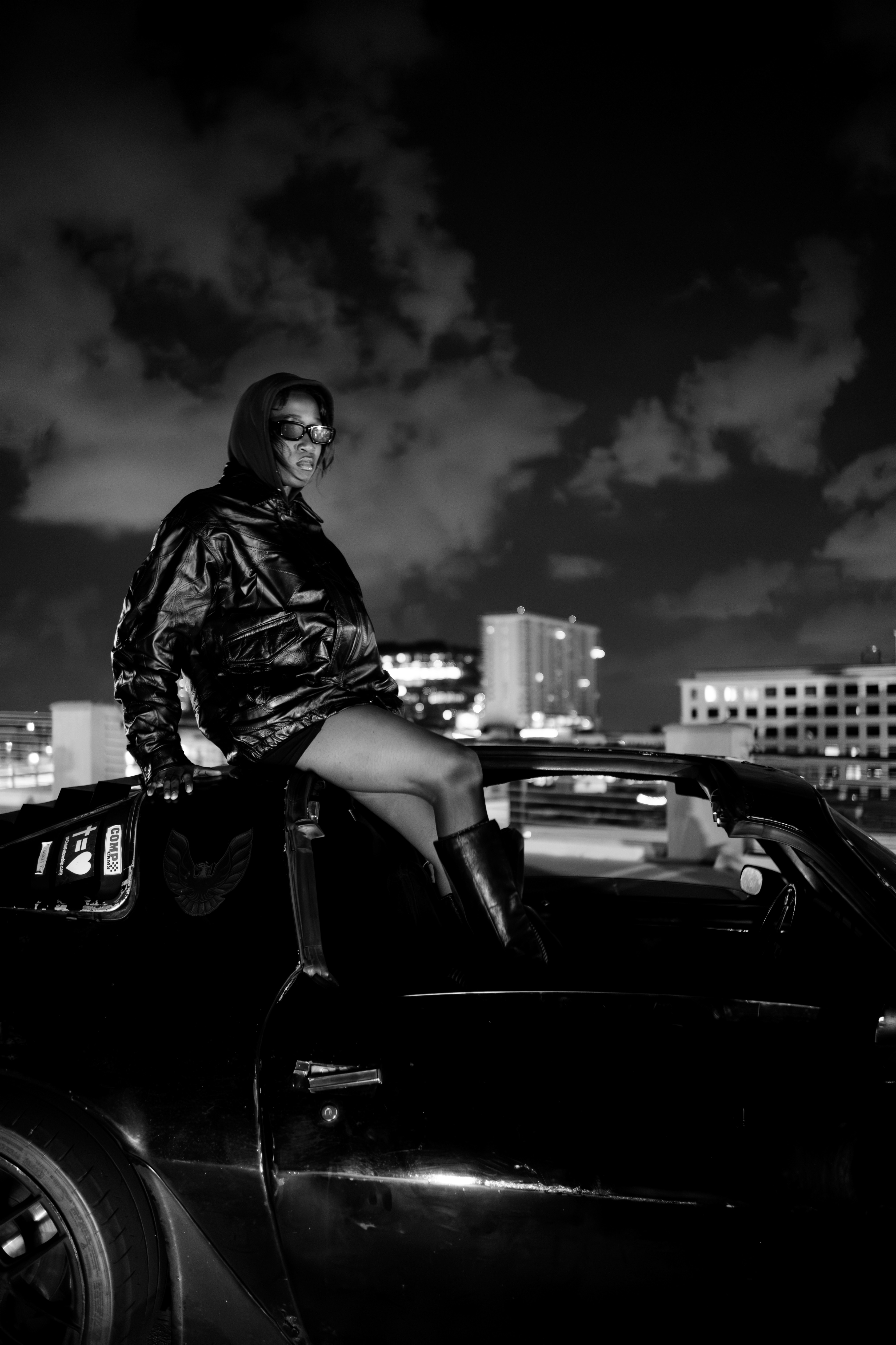Noir on Steel: Hooded Muse Perched on a Convertible Beneath City Night