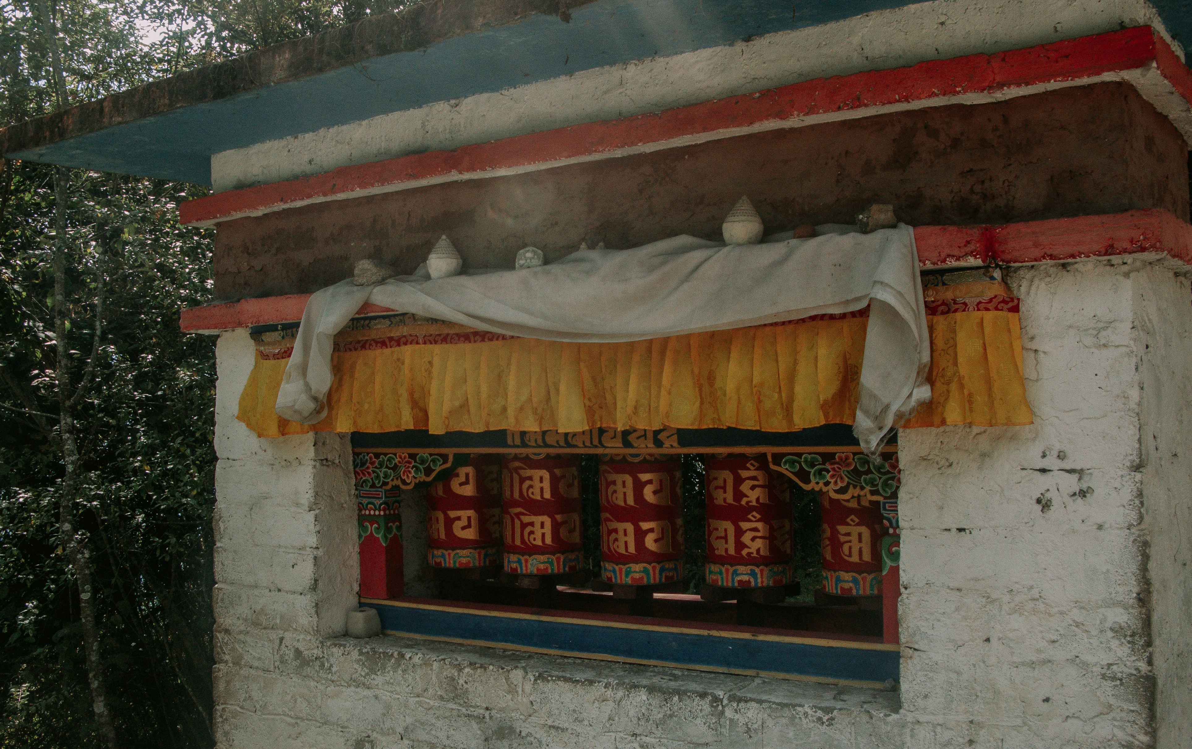 Small Tibetan shrine window housing red prayer wheels behind a yellow cloth canopy.