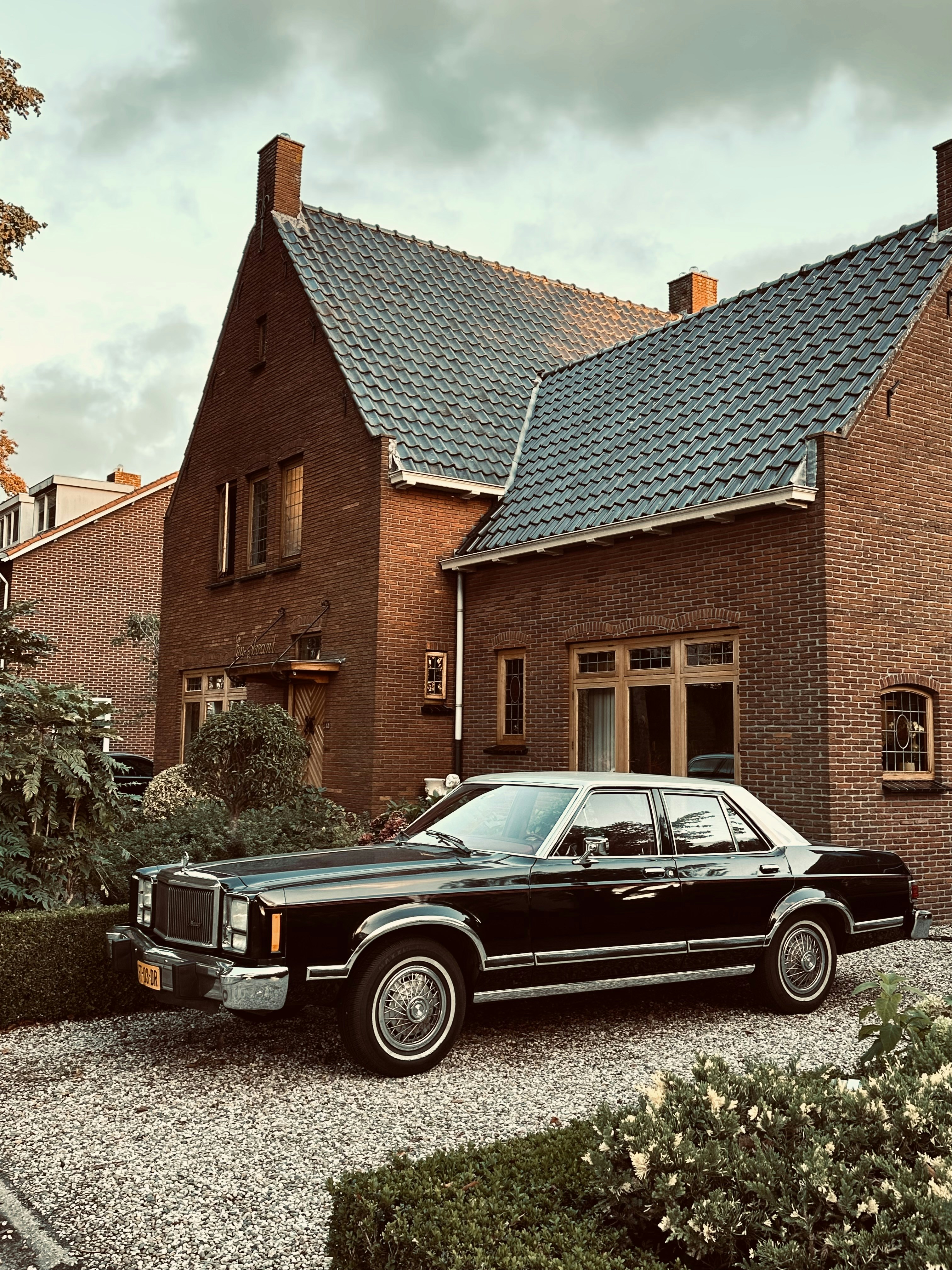 Classic black sedan parked in front of a charming brick house with unique architectural features, surrounded by lush greenery.