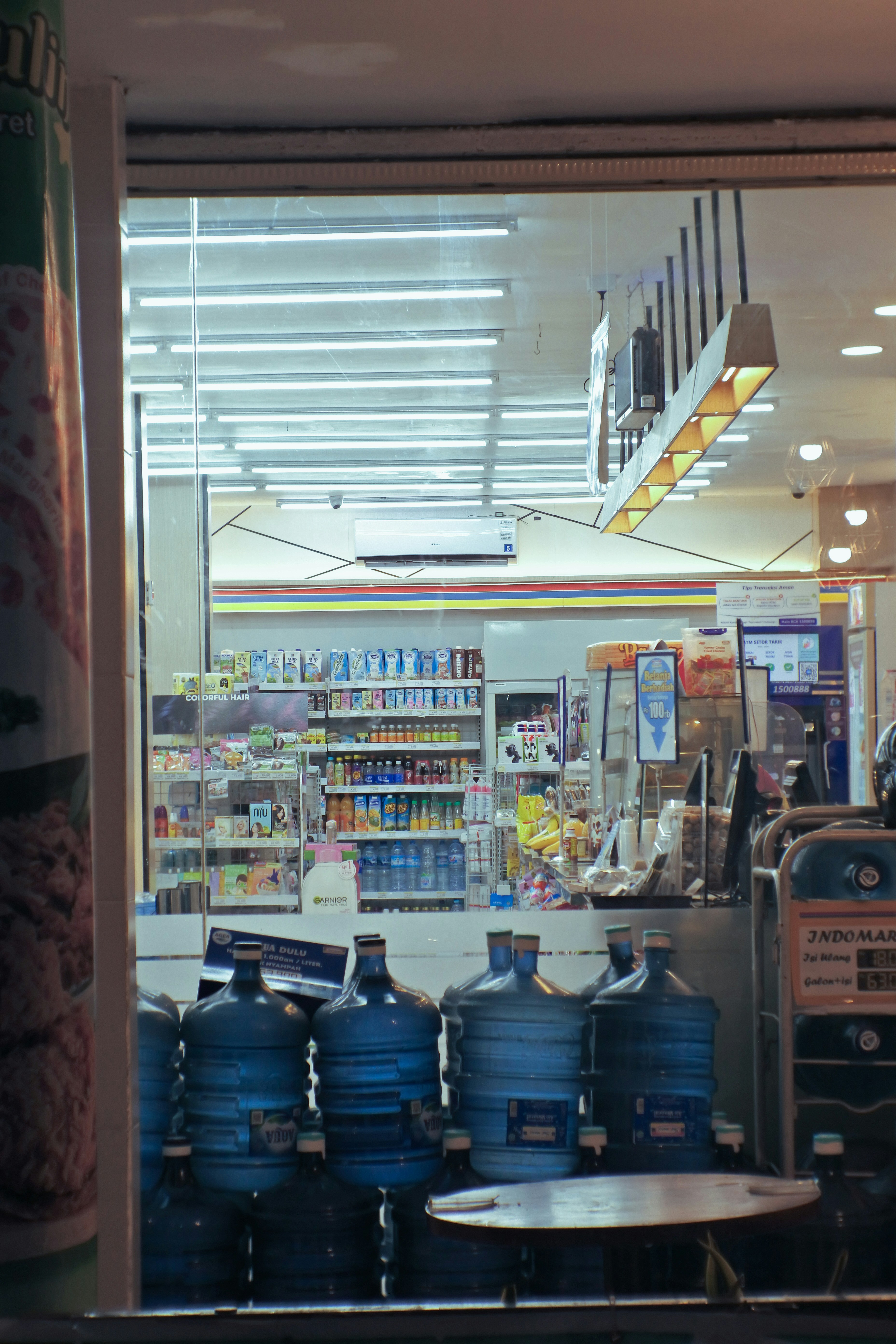 Store interior seen through a glass door with large blue water jugs in the foreground and well-lit shelves in the background.