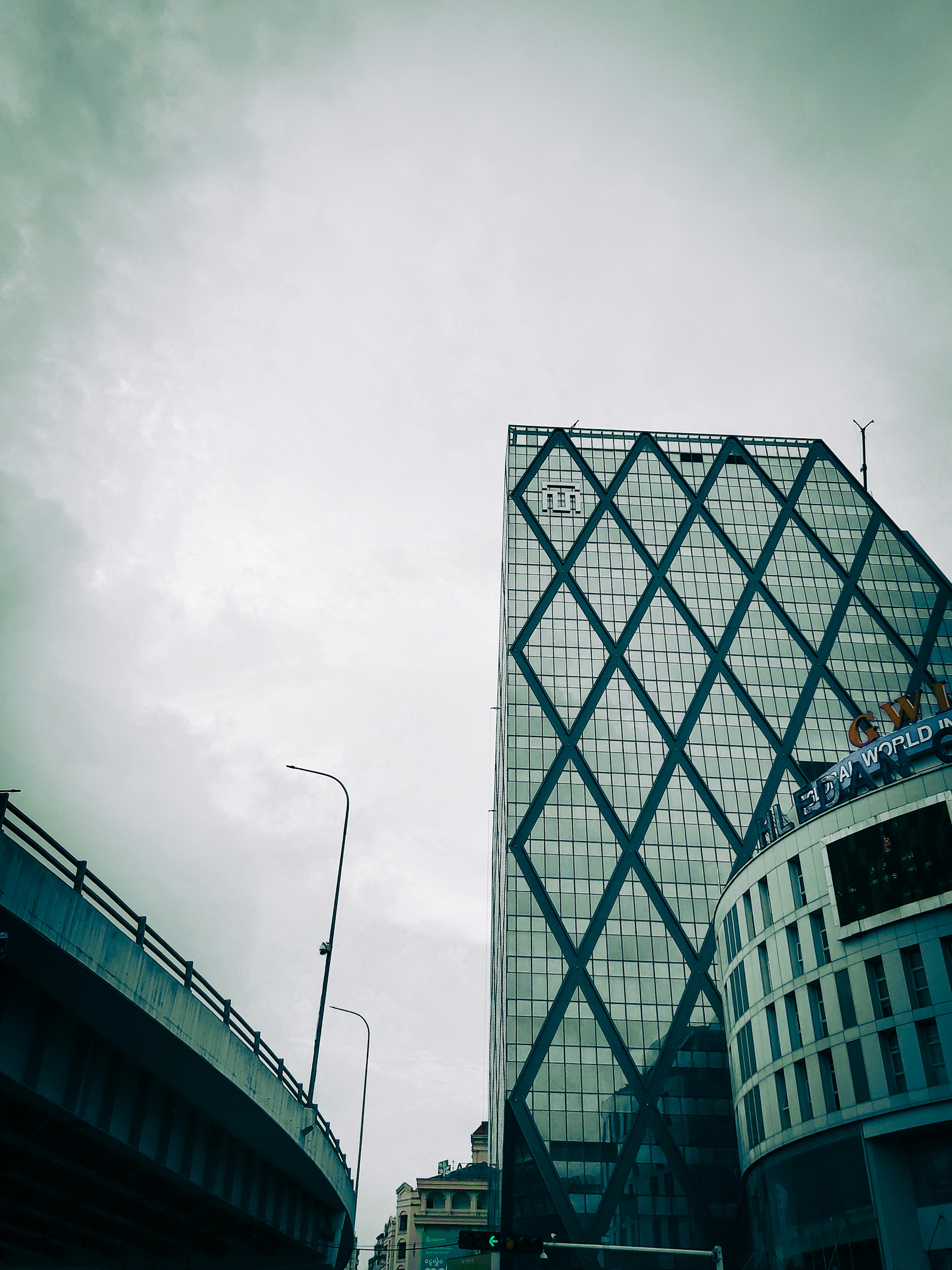 A modern glass tower with a blue diamond lattice façade rises beside a curved, white commercial building under a pale, overcast sky.