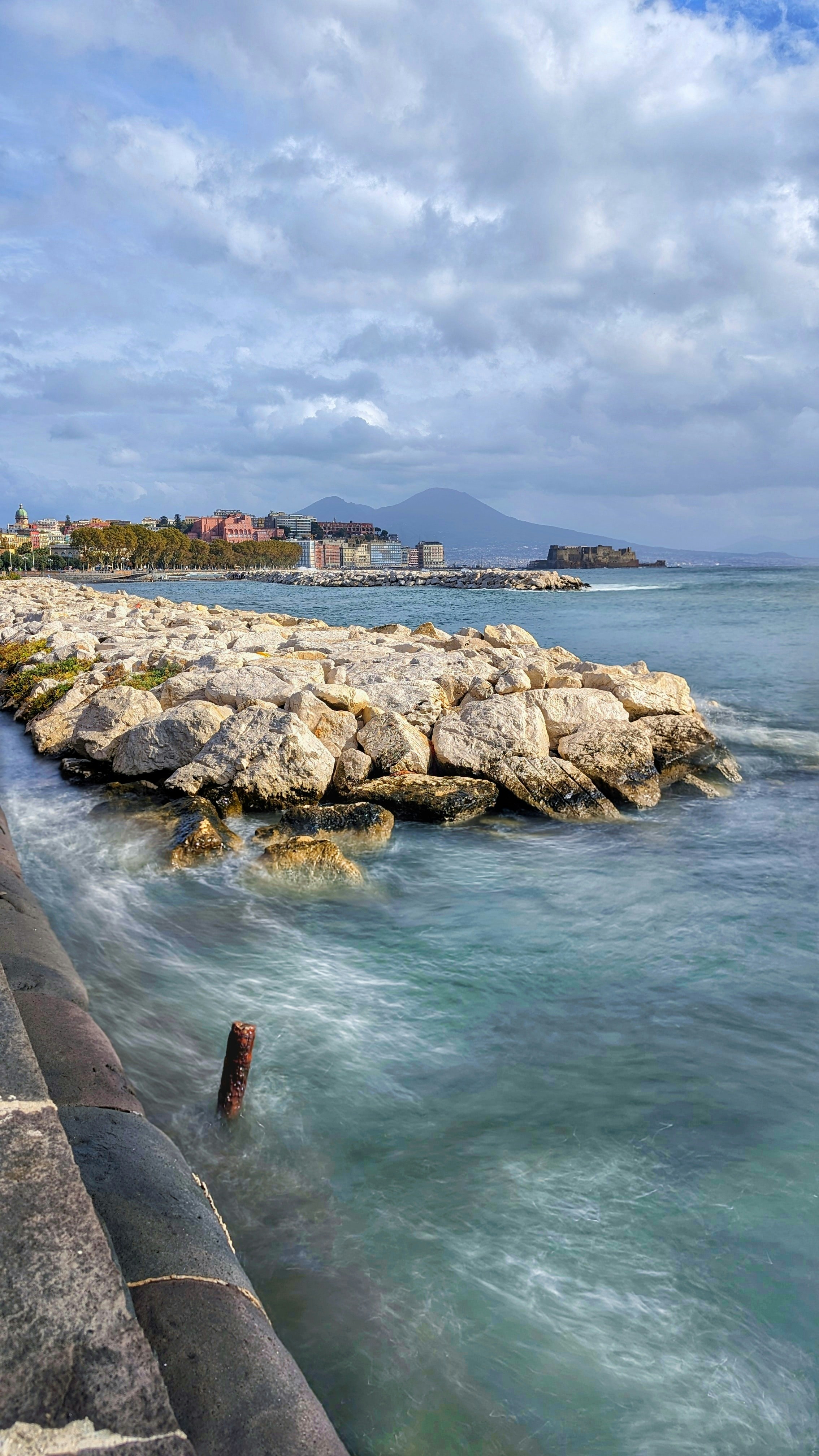 Coastal photograph of a rocky breakwater extending into turquoise water, with a town and distant mountains beneath a cloudy sky.