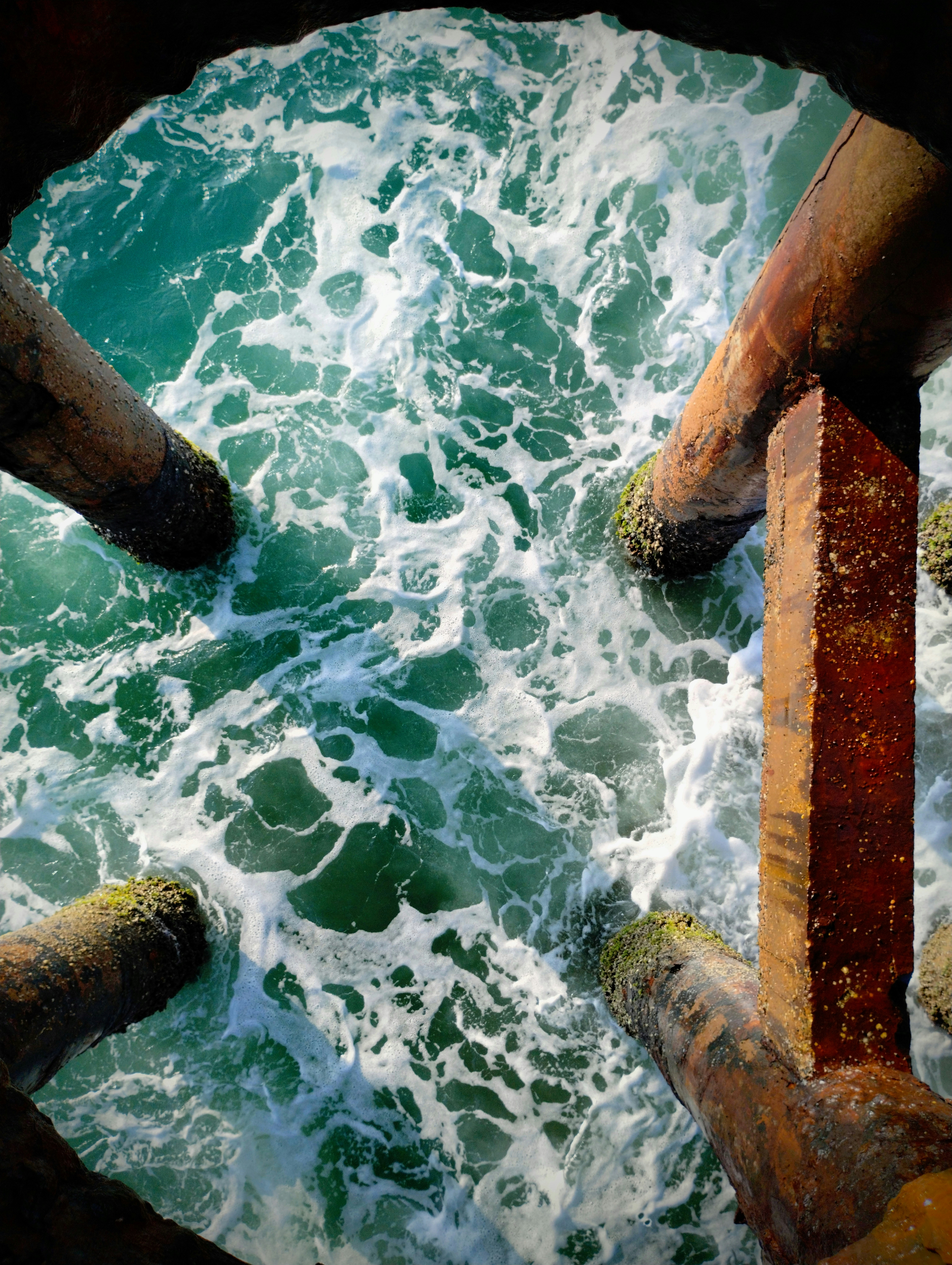 Turbulent turquoise water churns between gaps of rusted, weathered beams. The foamy white swirls form a dynamic pattern framed by the corroded metal.