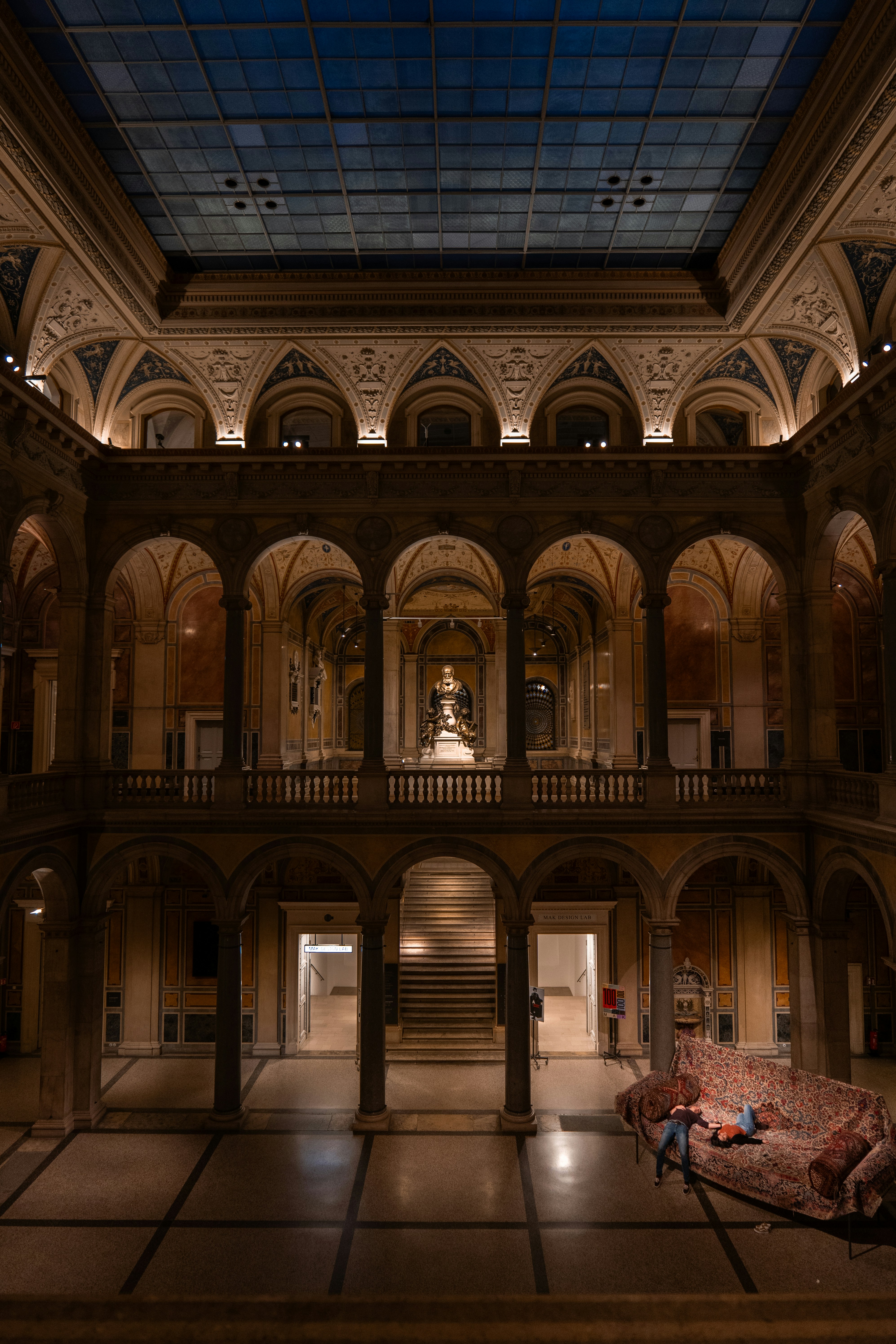 Elegant interior photograph of a grand arcaded hall with a central statue illuminated by warm lighting beneath a glass skylight.