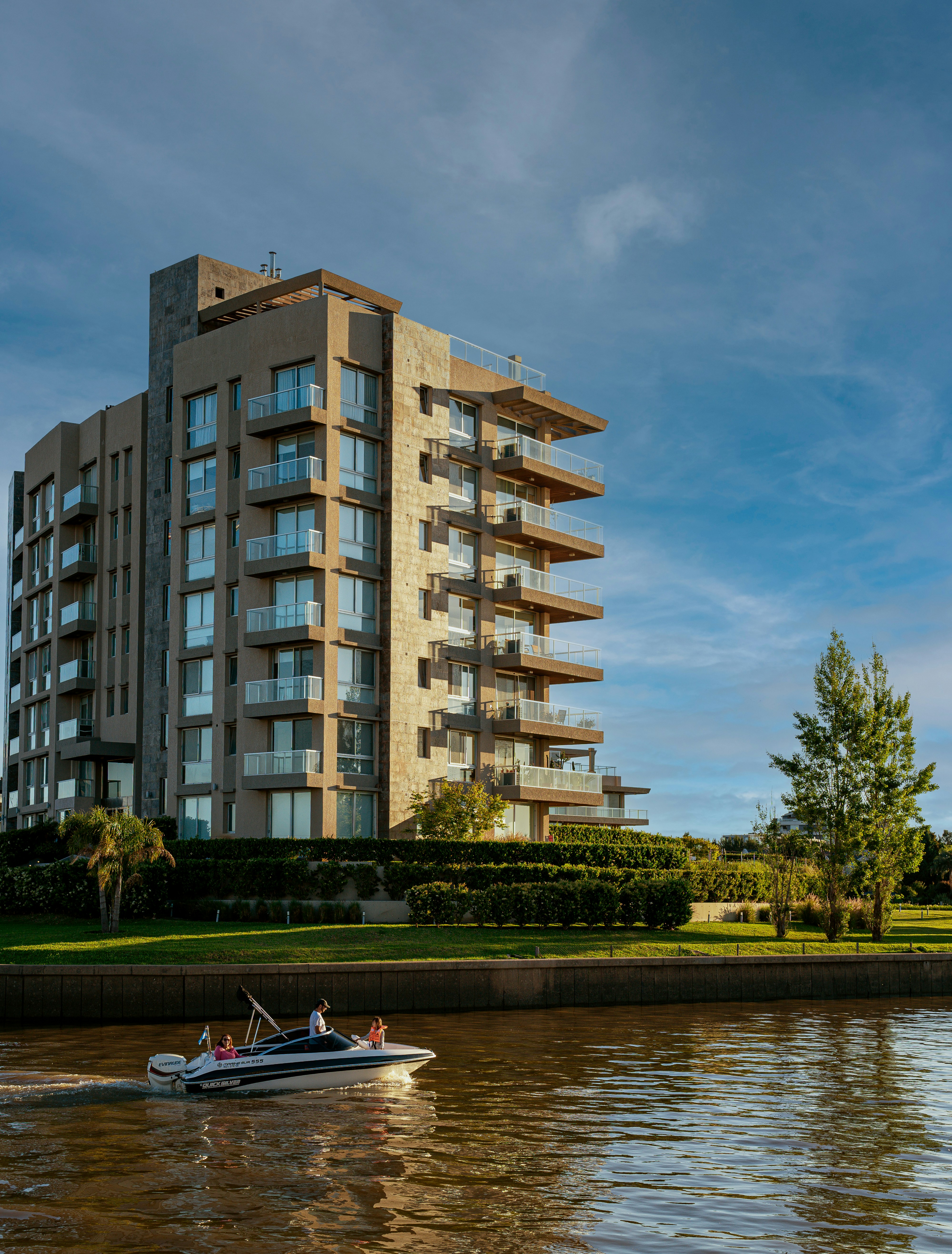 Modern apartment building alongside a tranquil river, with a boat gliding by and lush greenery in the foreground.
