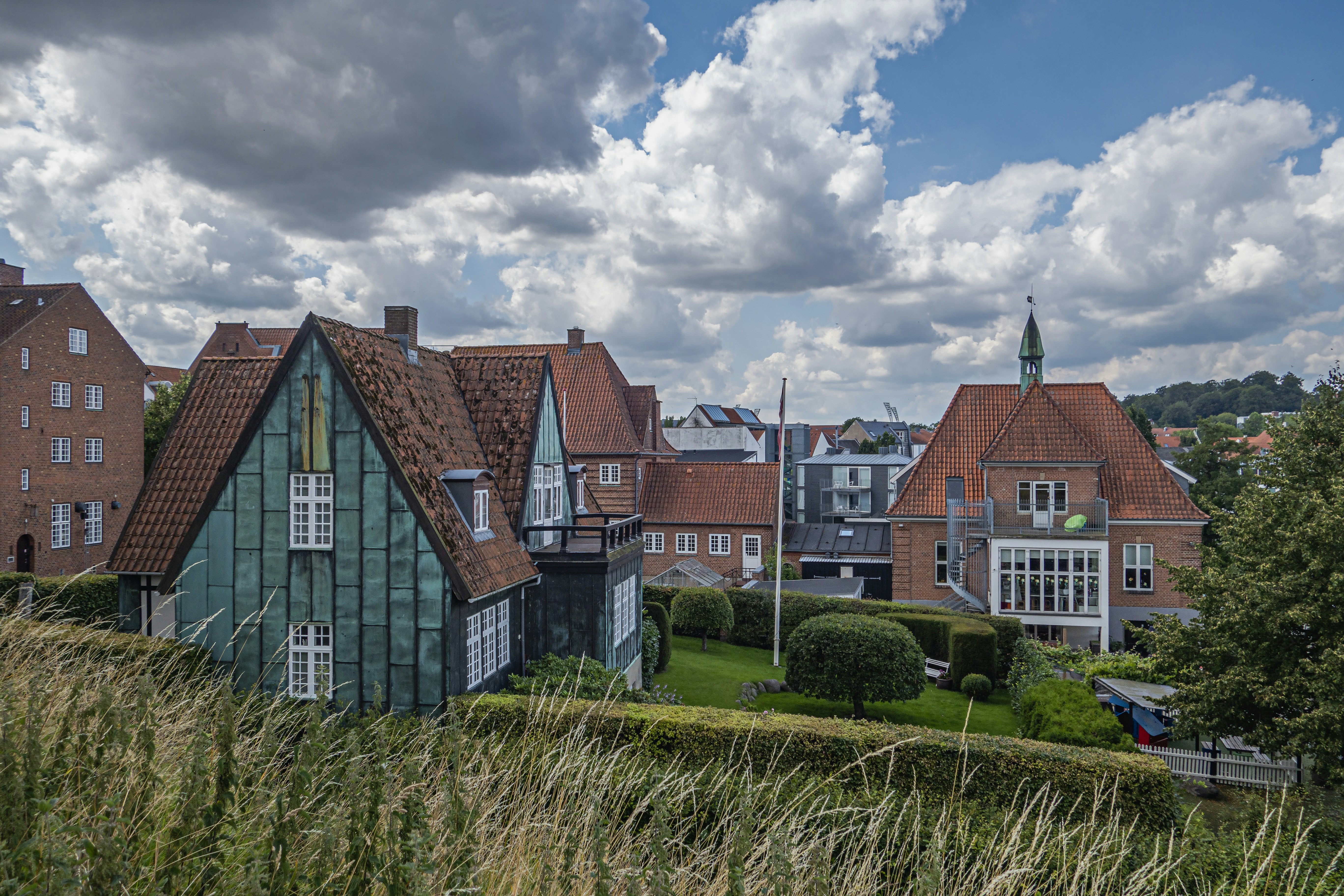 Row of quaint houses with steep roofs set against a backdrop of lush greenery and billowing clouds.