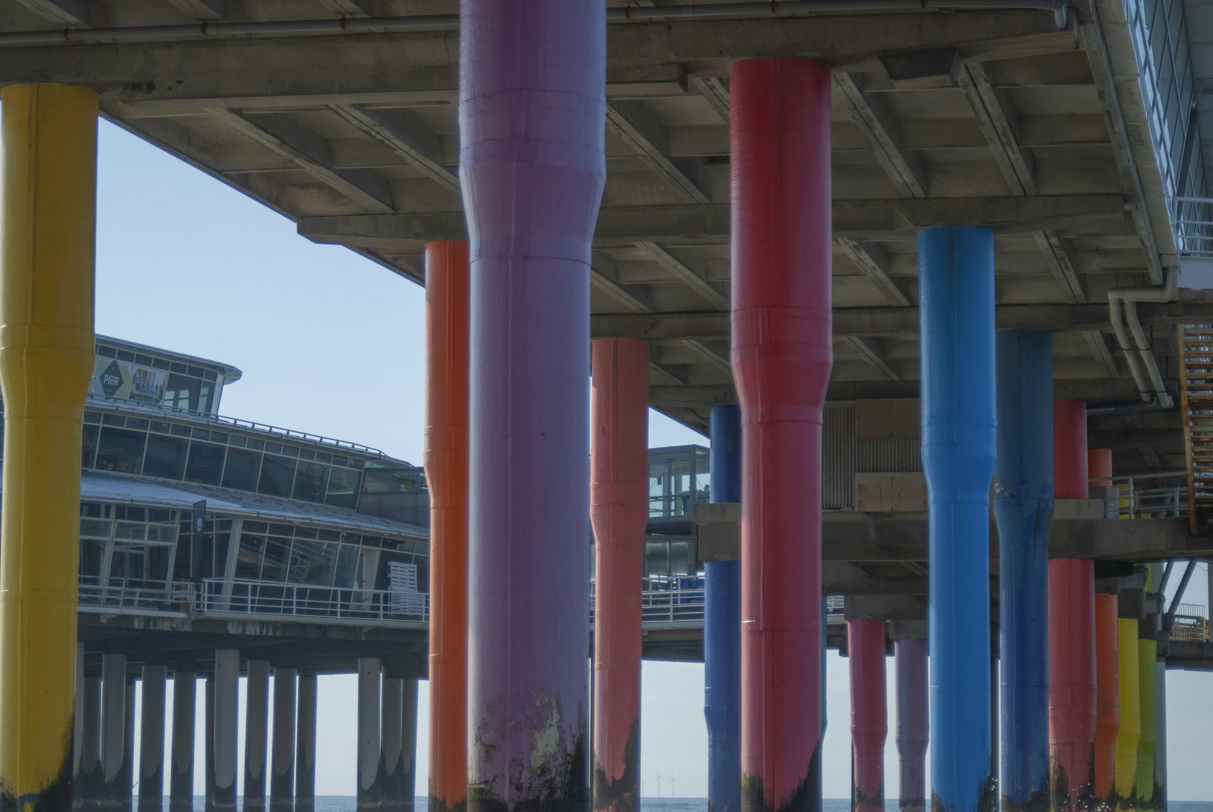 Colorful pillars supporting a pier, showcasing a blend of bright hues against a clear sky. The architectural structure creates a dynamic interplay of color and form.