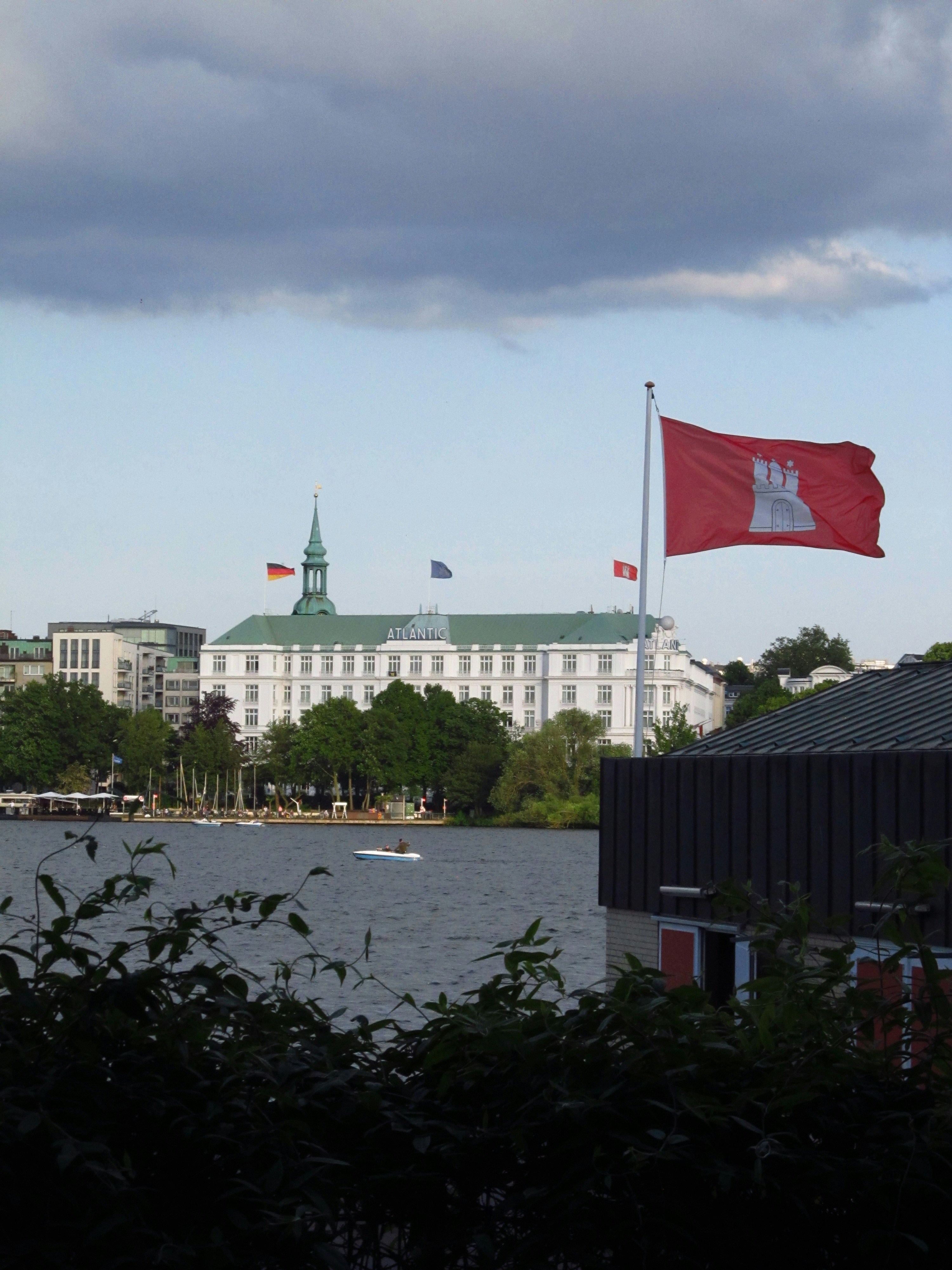 Riverside cityscape across calm water featuring a white building with a green roof. A tall red flag on a pole dominates the foreground, with leafy brush along the shore.