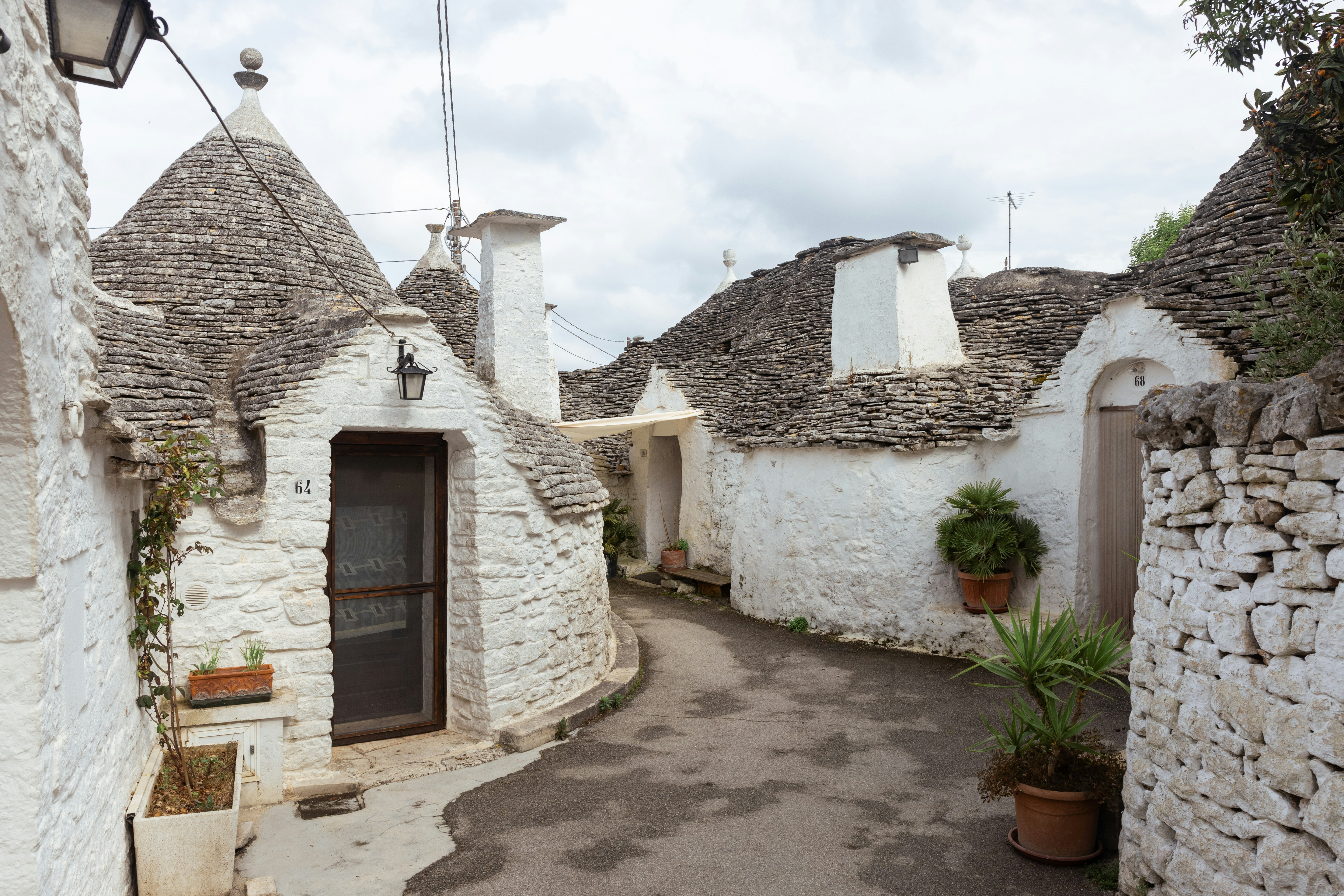 Cobblestone street with white stone trulli featuring conical roofs under a cloudy sky.