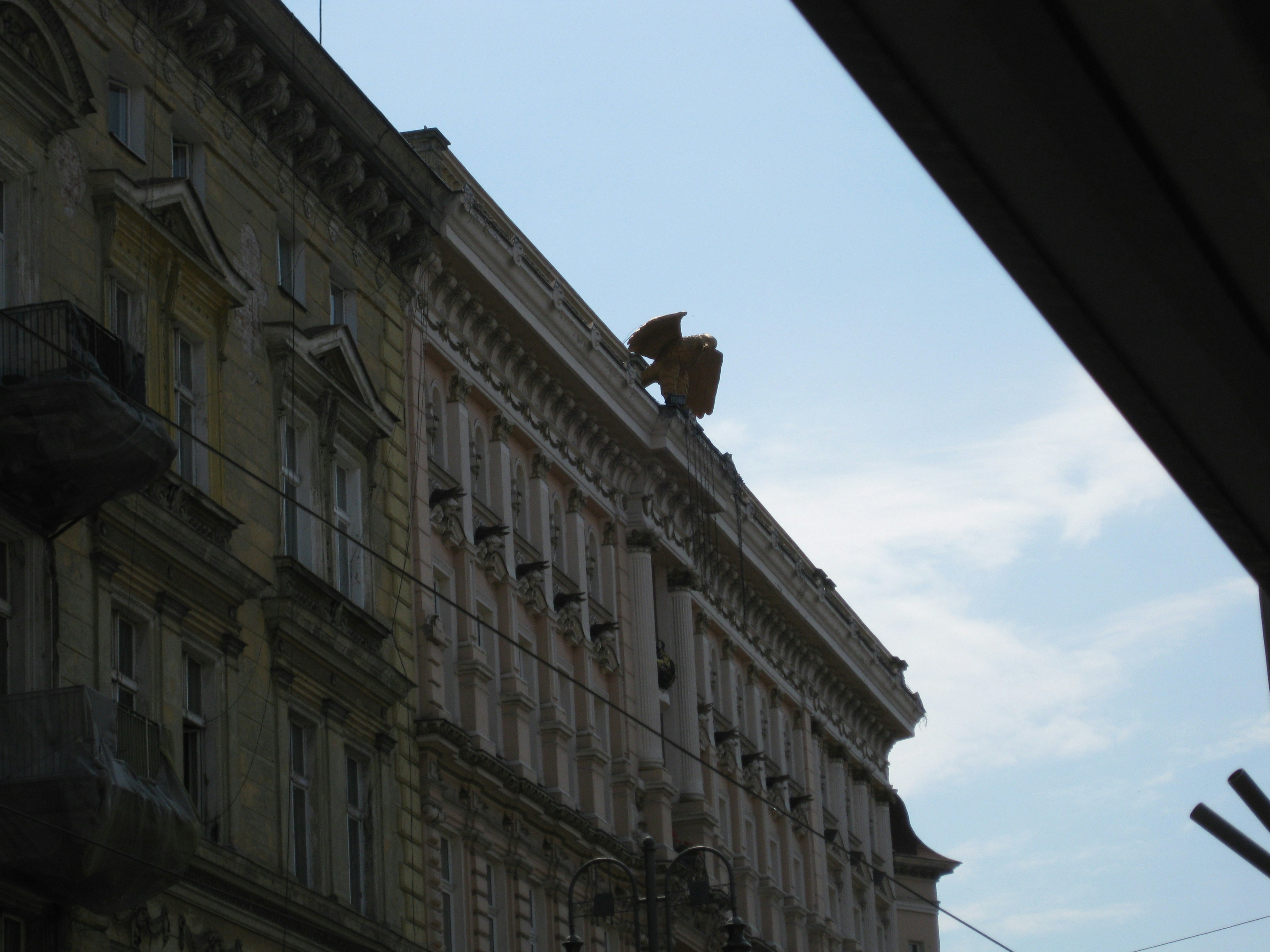 Ornate multi-story building facade with a sculpted statue atop the roof, viewed from a street angle against a pale blue sky.