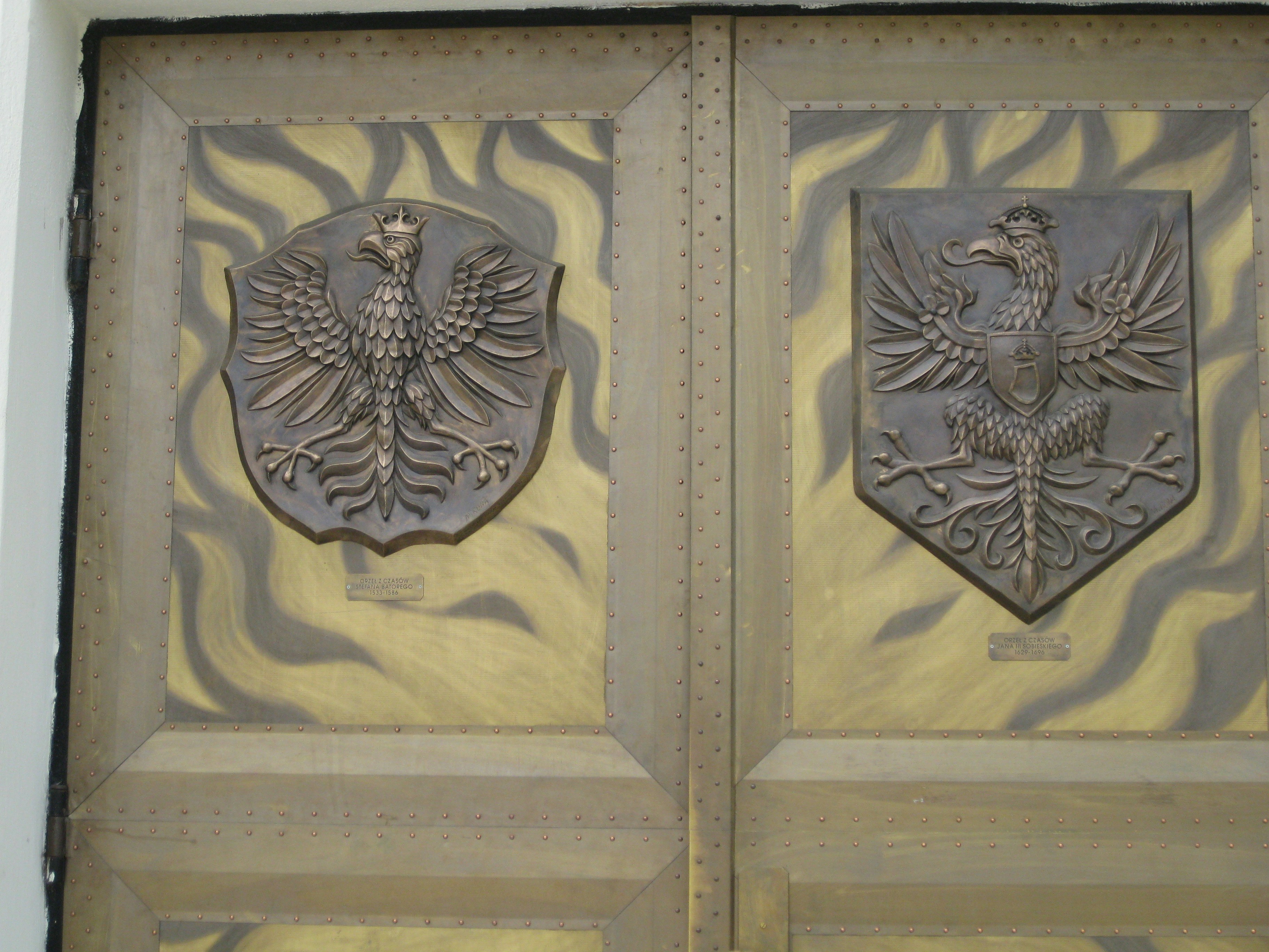 Two bronze doors feature embossed heraldic shields—eagle on the left and dragon on the right—set against a textured, wavy background.