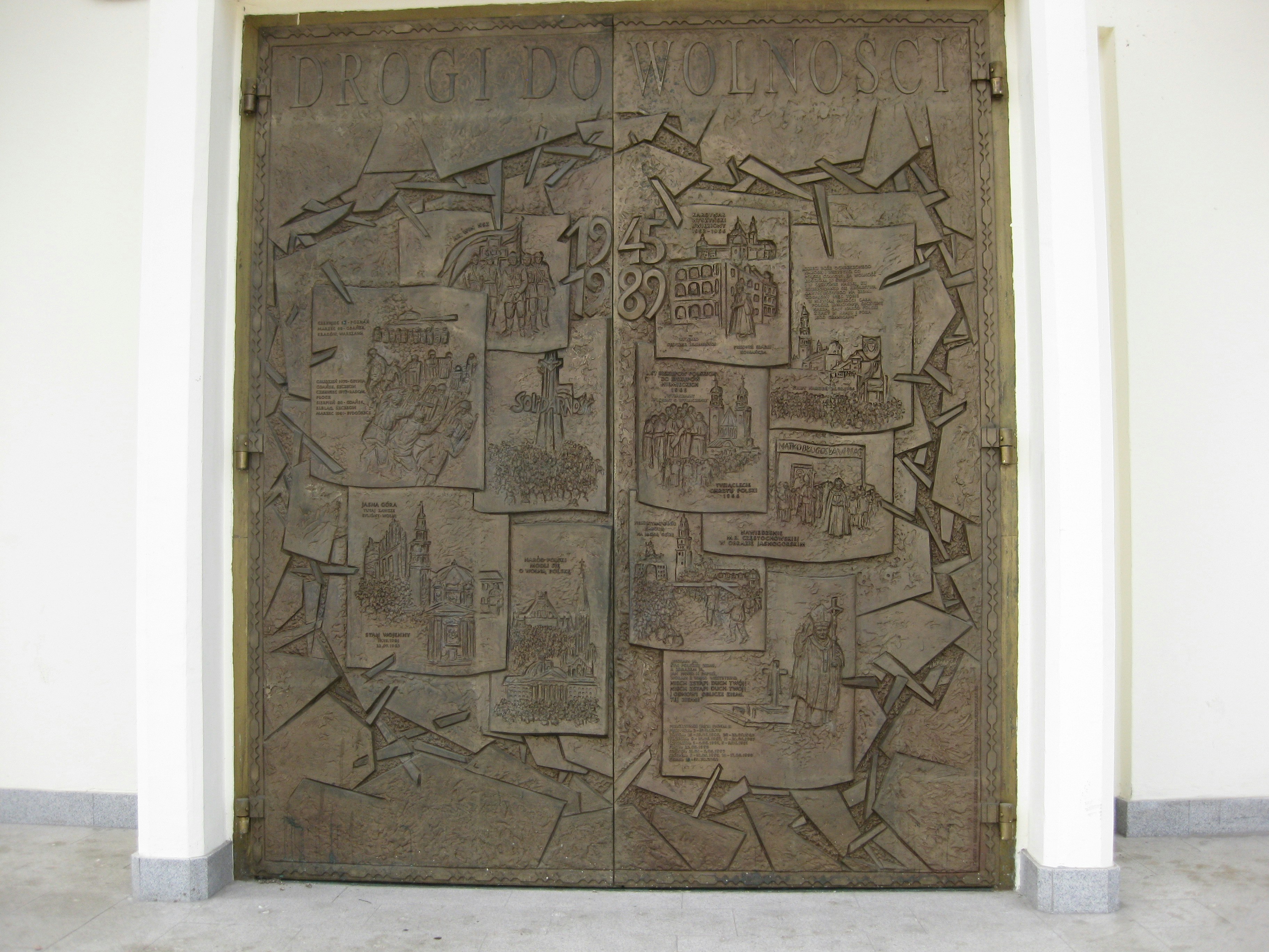 Heavy bronze door carved with panels and jagged crack motifs frames a collage of inscriptions. This doorway guards a historical display.