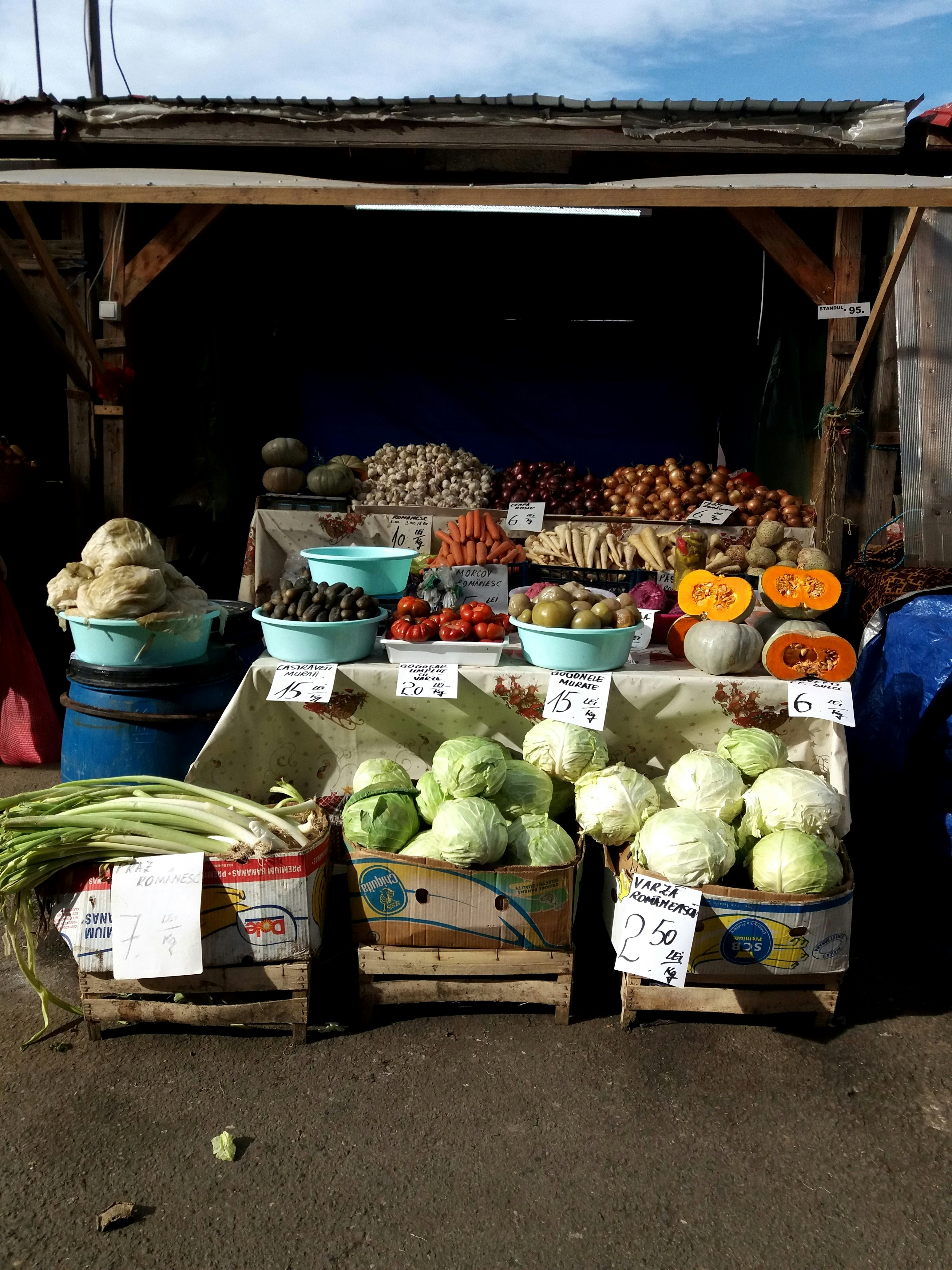 Wooden market stall displaying an array of fresh vegetables, including cabbages, pumpkins, and potatoes, under a sunny sky.