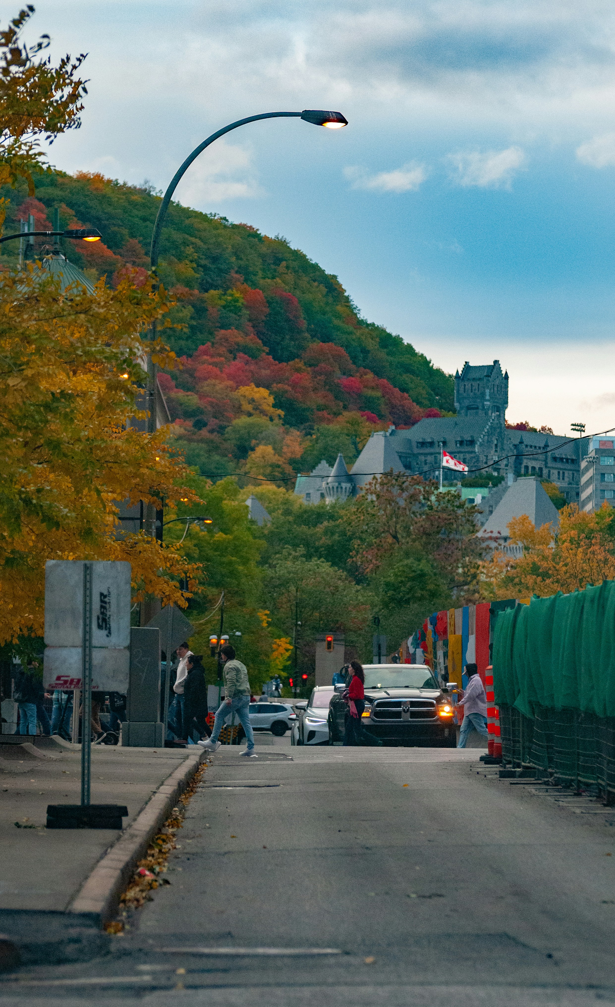 A city street lined with fall foliage and a distant castle-like building, with pedestrians and a vehicle near a construction barrier.
