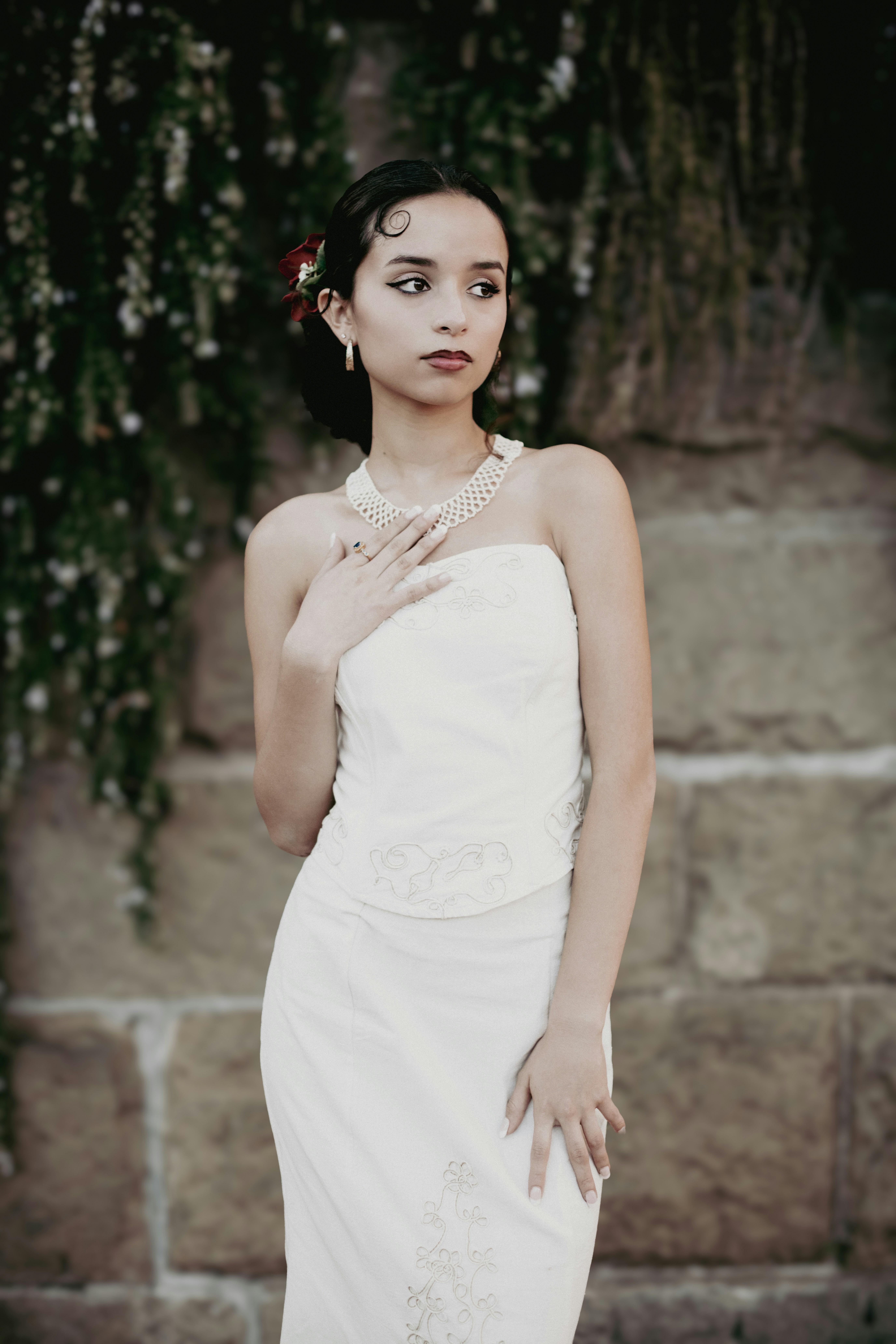 Woman in a white dress standing against a stone wall with cascading greenery.