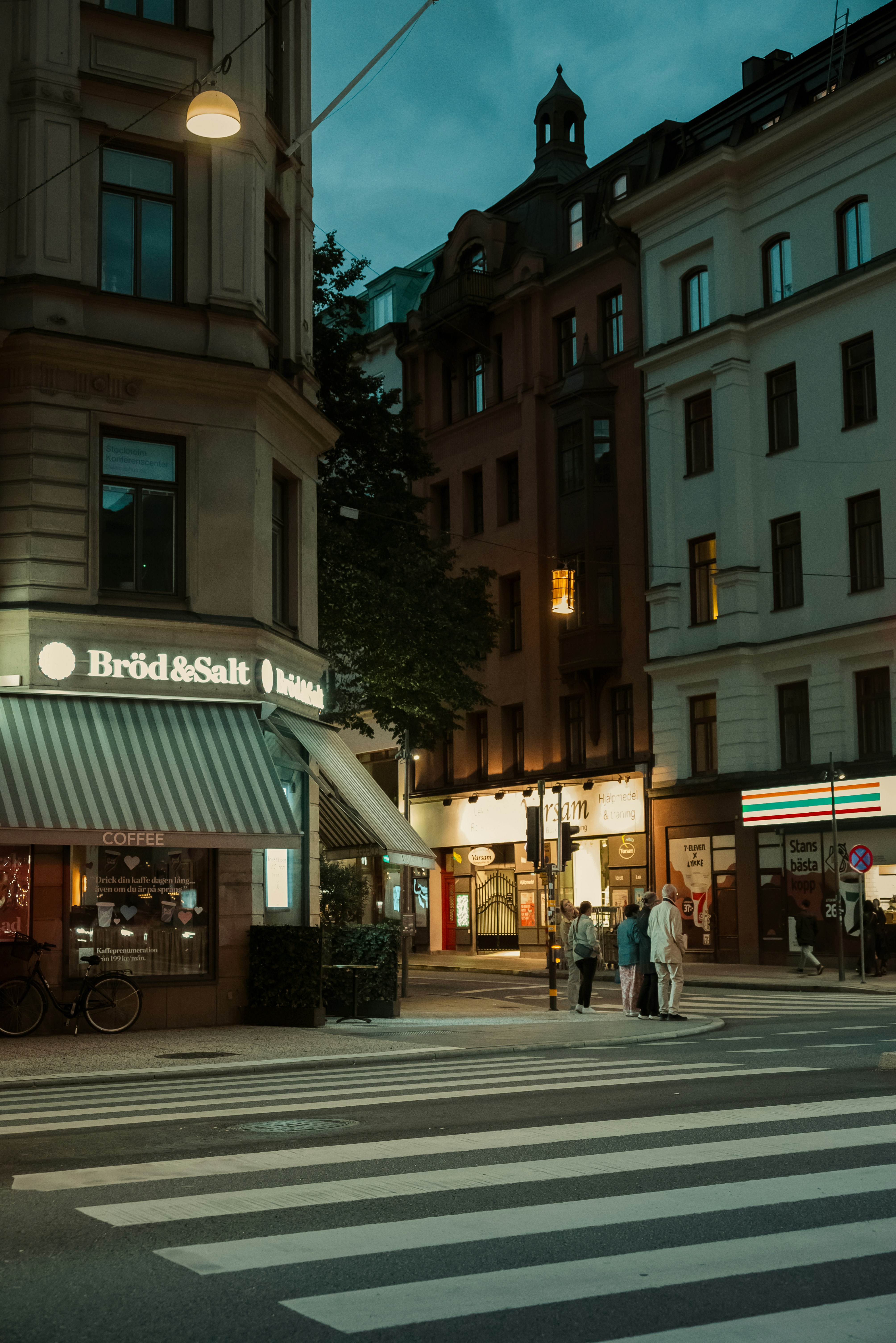 Dusk Crossing by Bröd&Salt: Pedestrians beneath amber storefronts along a quiet European street.