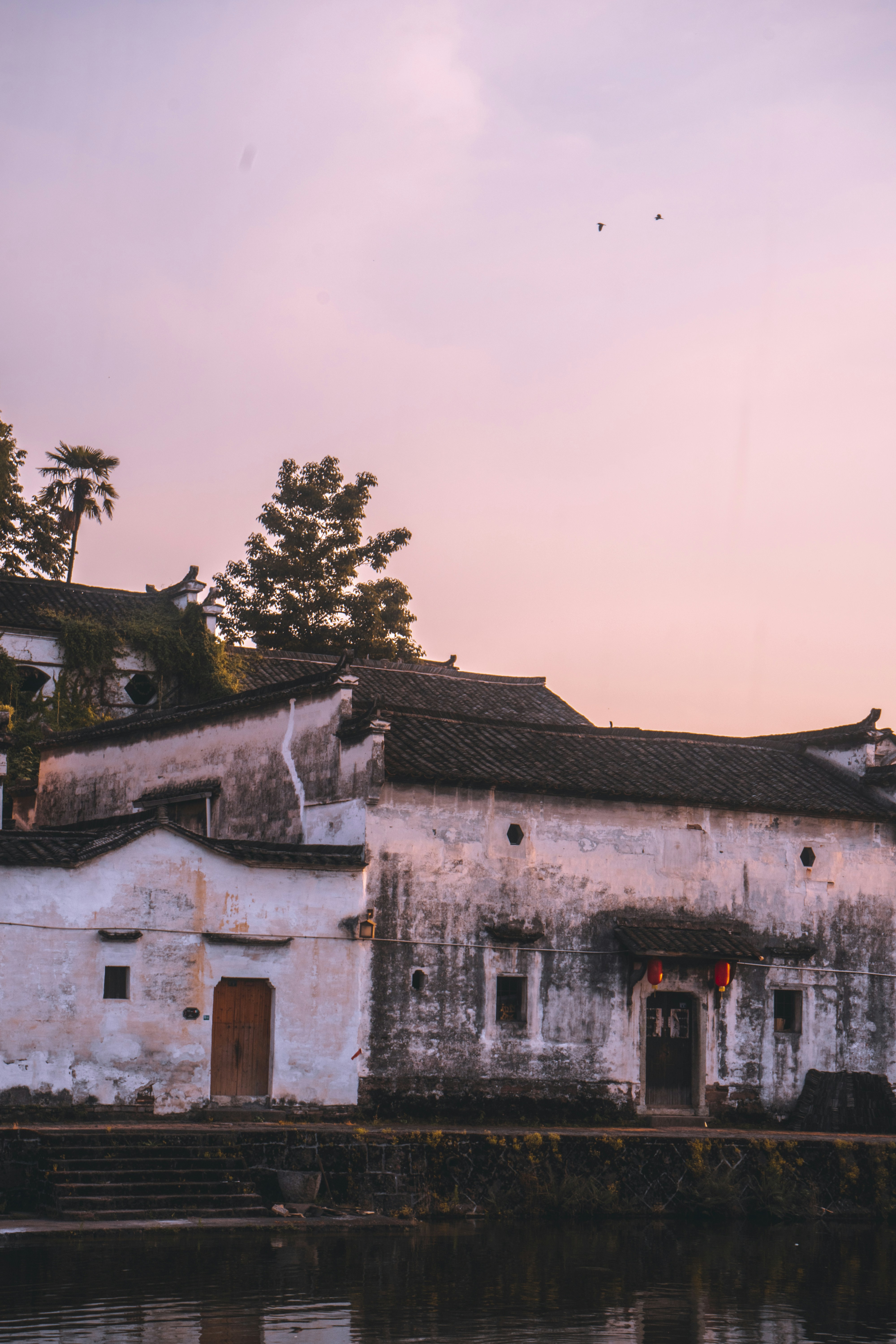 Historic white building with weathered walls beside a tranquil river under a soft pink sky.