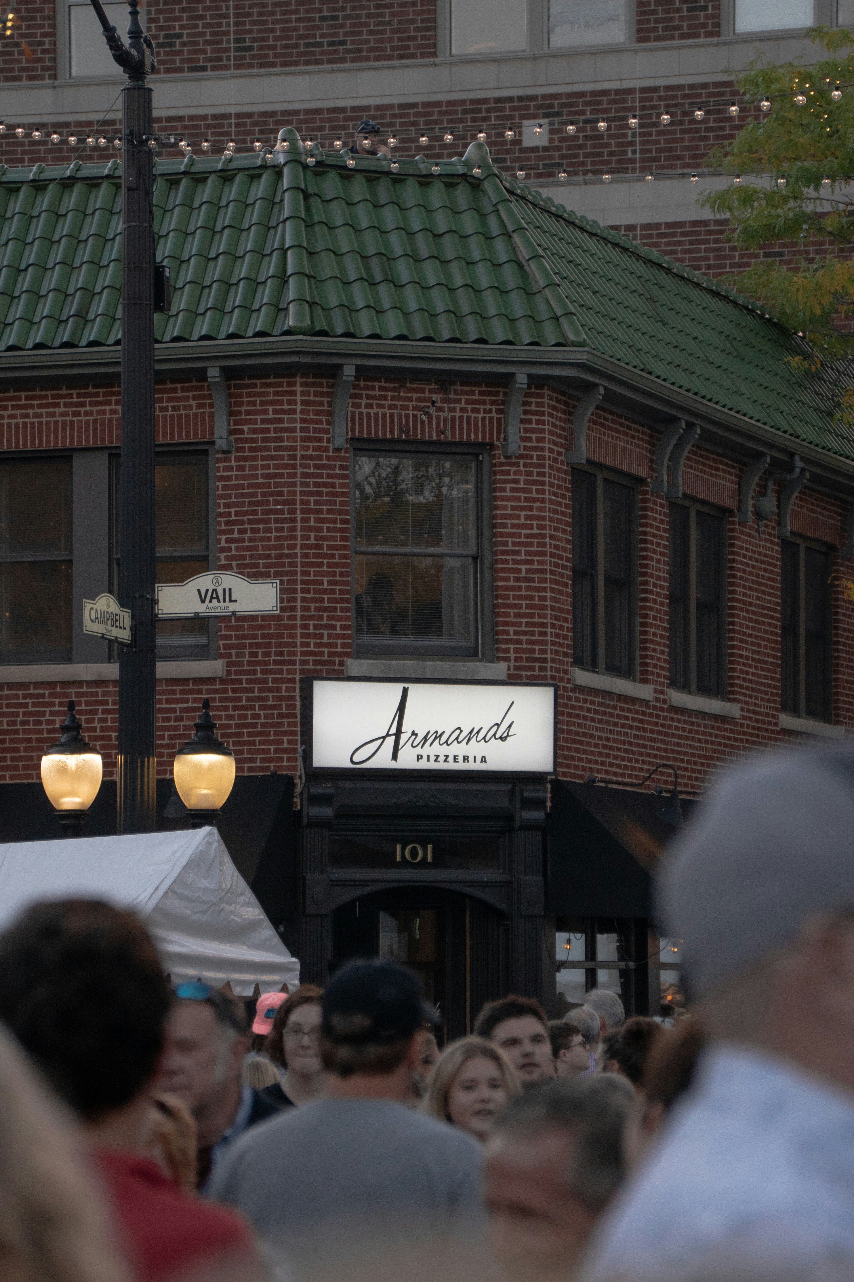 Crowd gathered in front of a brick corner building with a green tiled roof and illuminated signs.