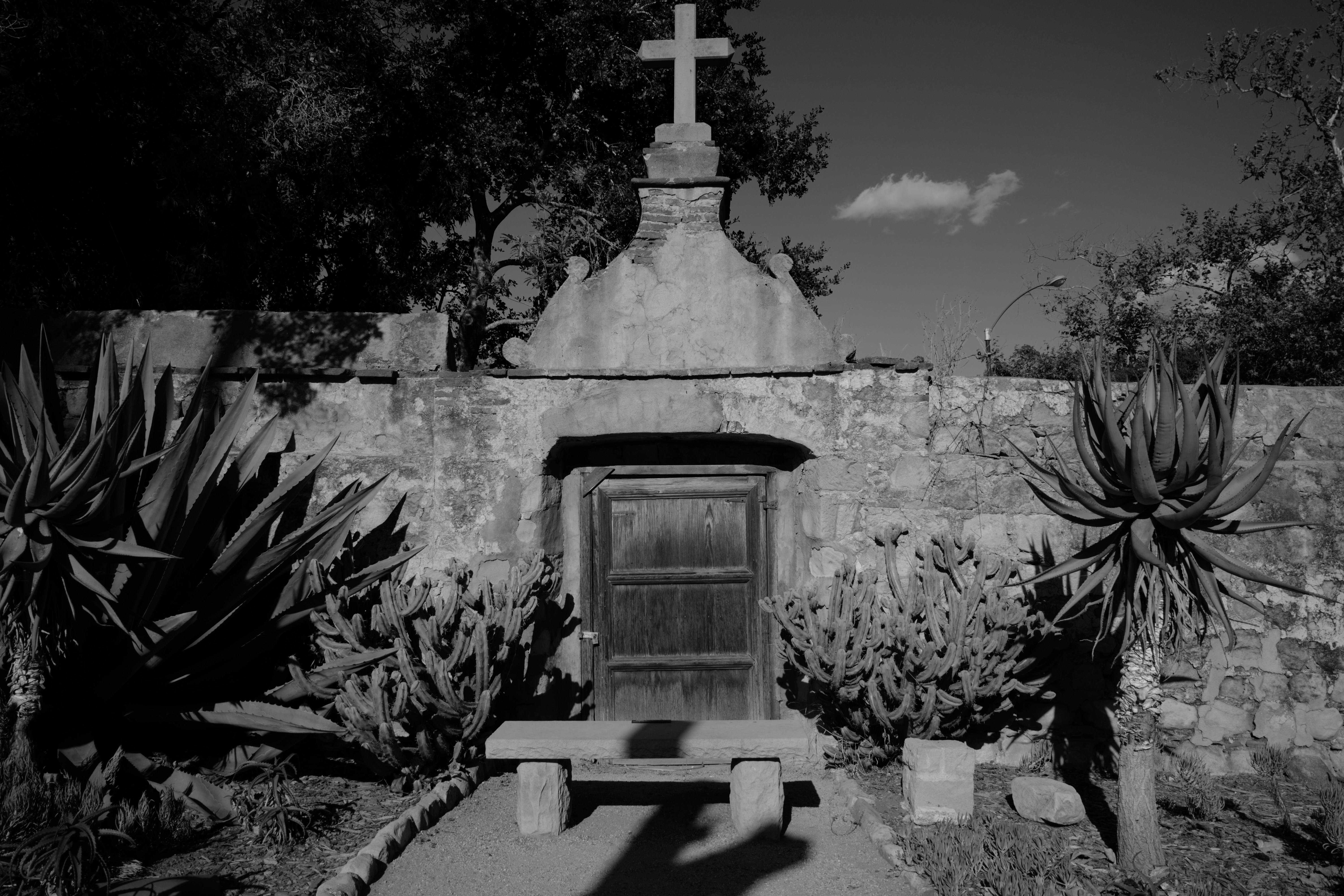 Grayscale photograph of a weathered stone gate and doorway crowned with a cross, set in a sunlit courtyard.
