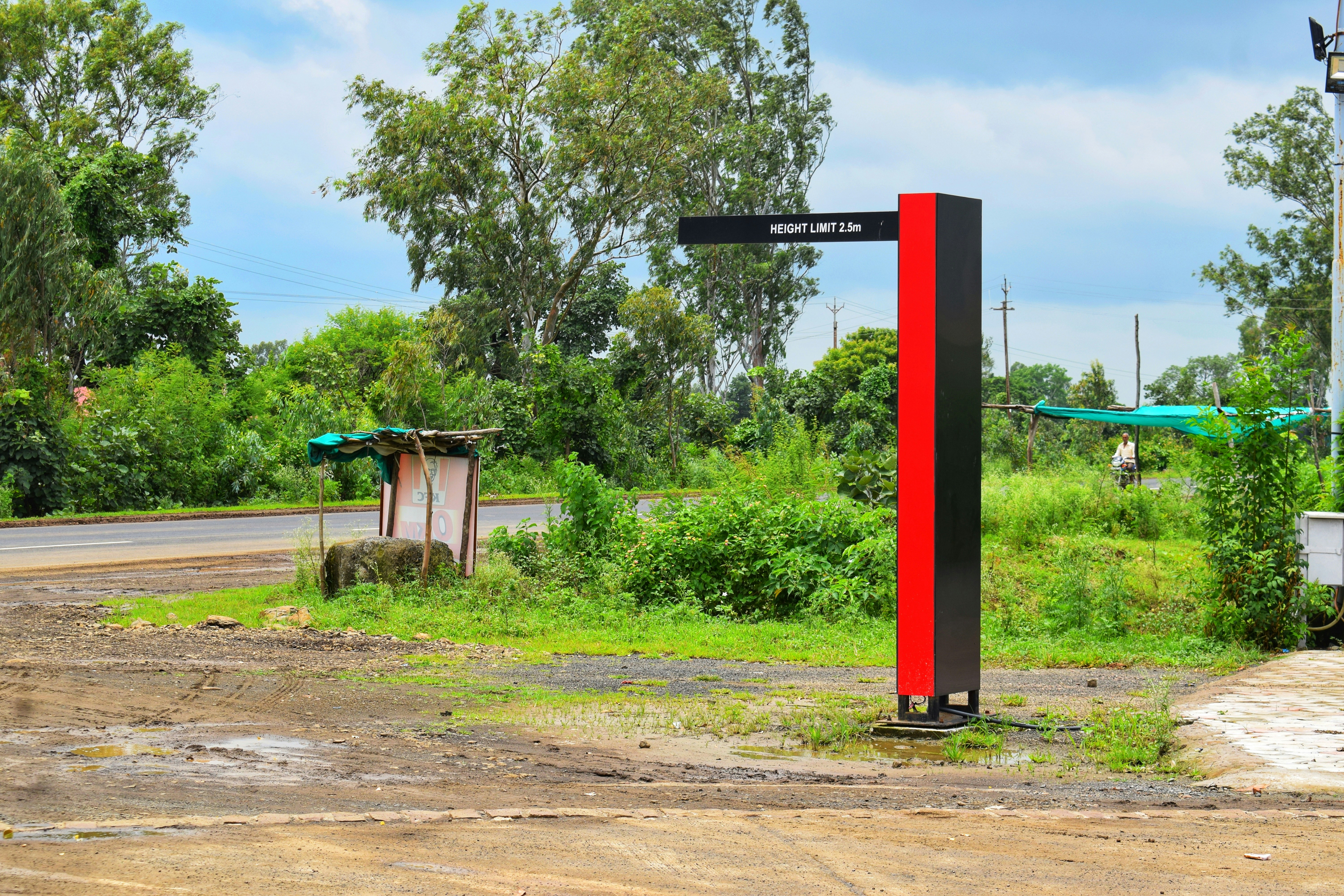 A red-and-black height-limit column rises beside a muddy roadside, surrounded by green shrubs and trees.