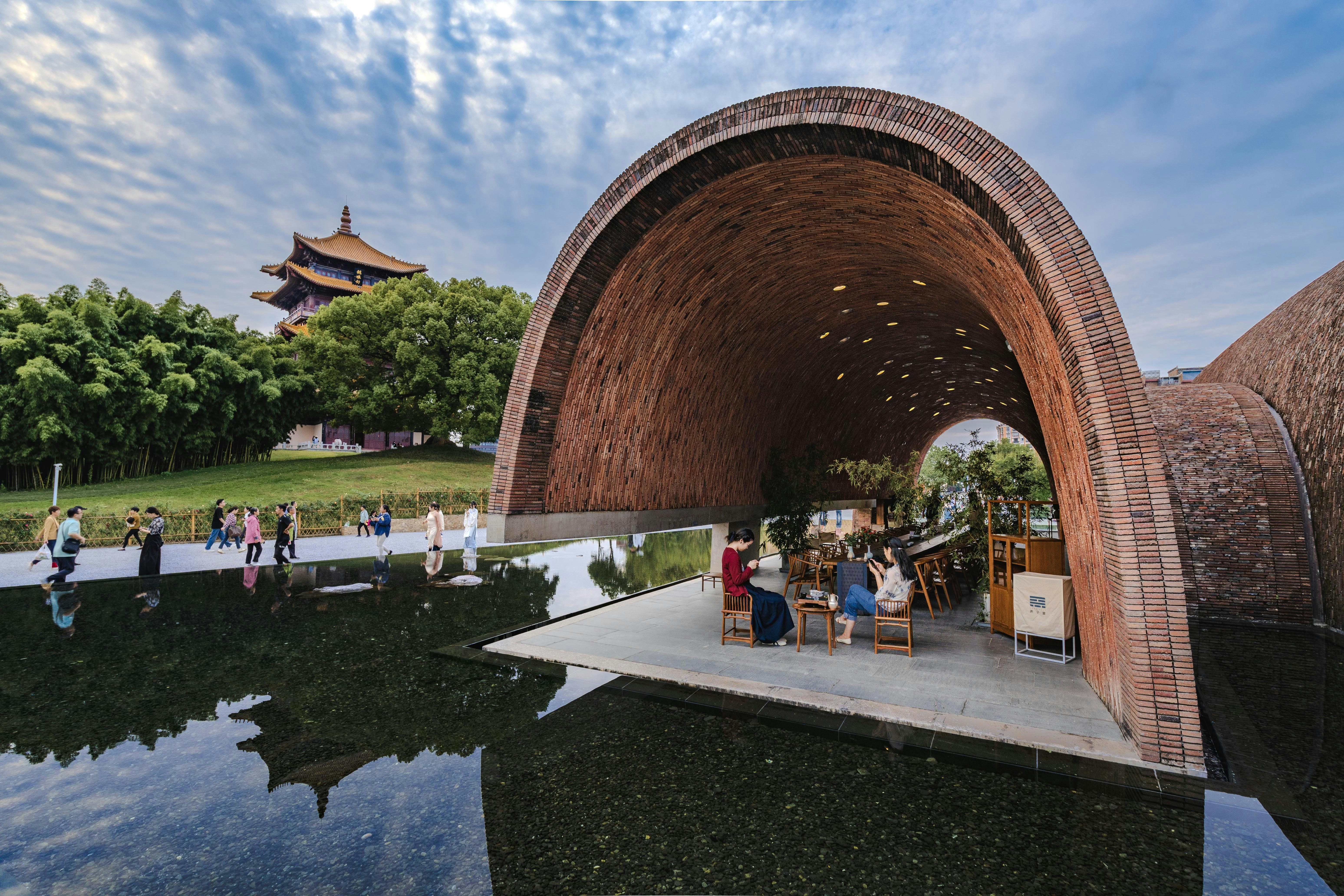 Curved brick structure reflecting in tranquil water with visitors nearby, set against a backdrop of lush greenery and a traditional pagoda.