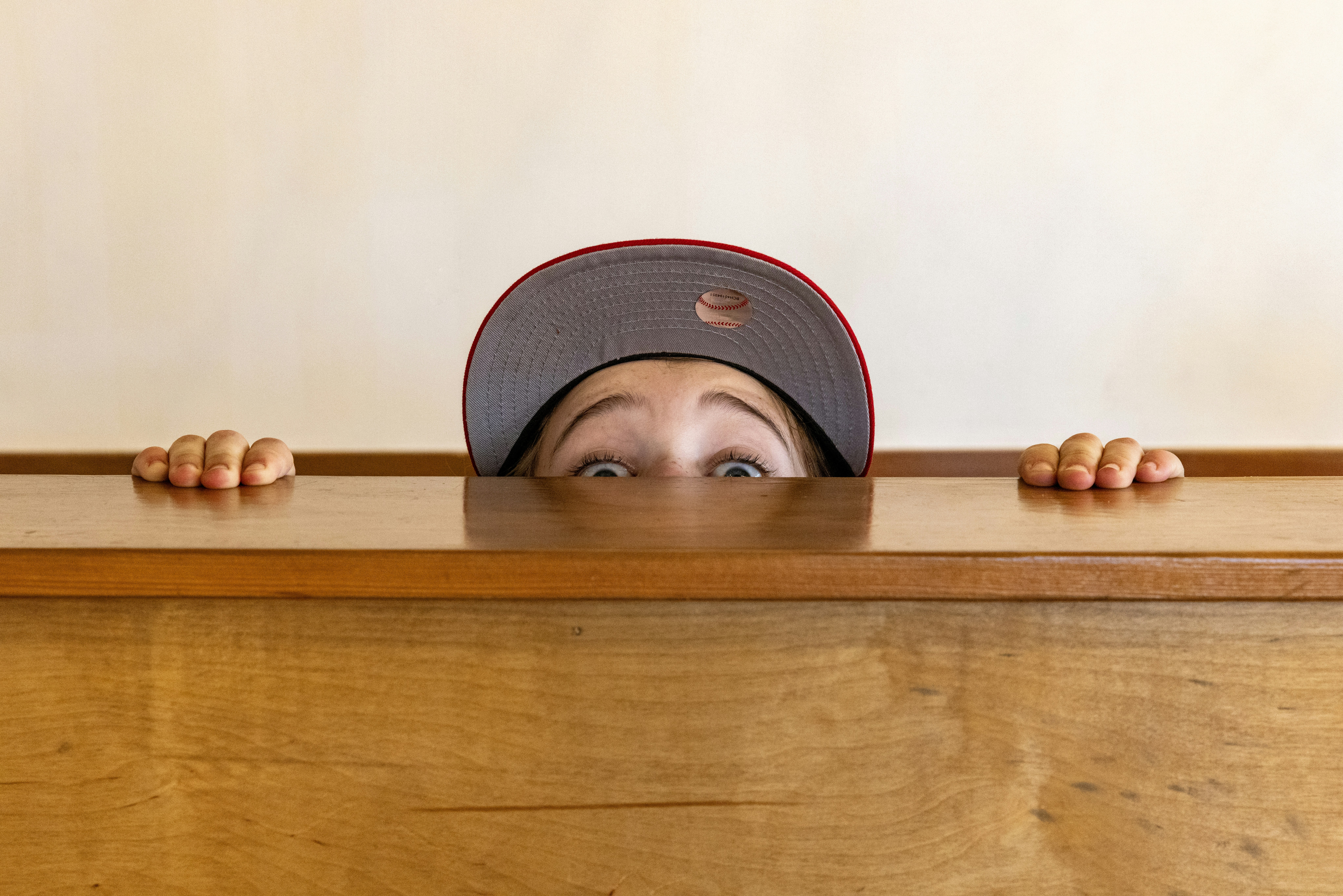 A child peeking over a wooden barrier, eyes wide with curiosity, wearing a cap. The background is softly blurred, emphasizing the child's expression.