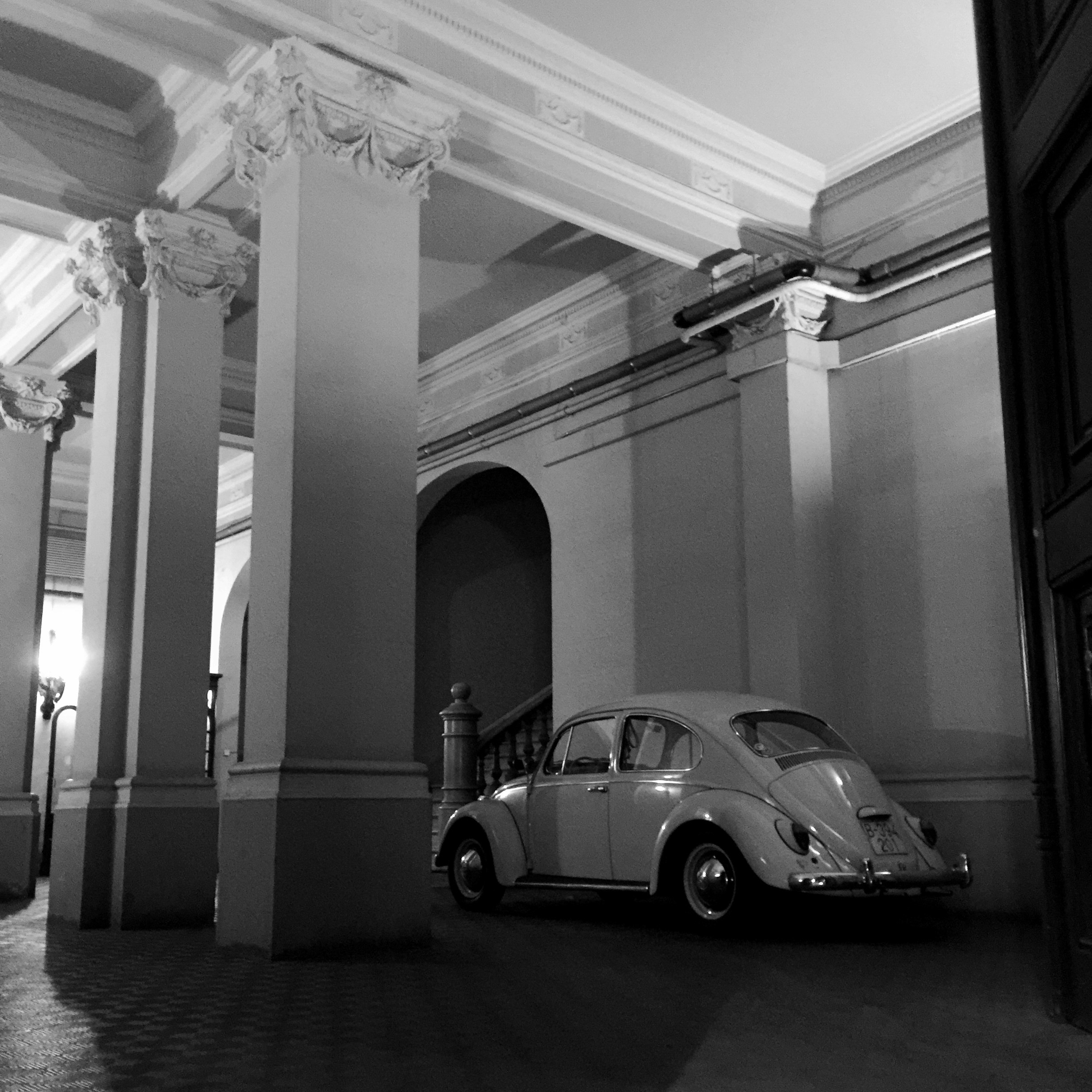 Monochrome photograph of a vintage Volkswagen Beetle parked beside tall Corinthian columns inside a grand interior.