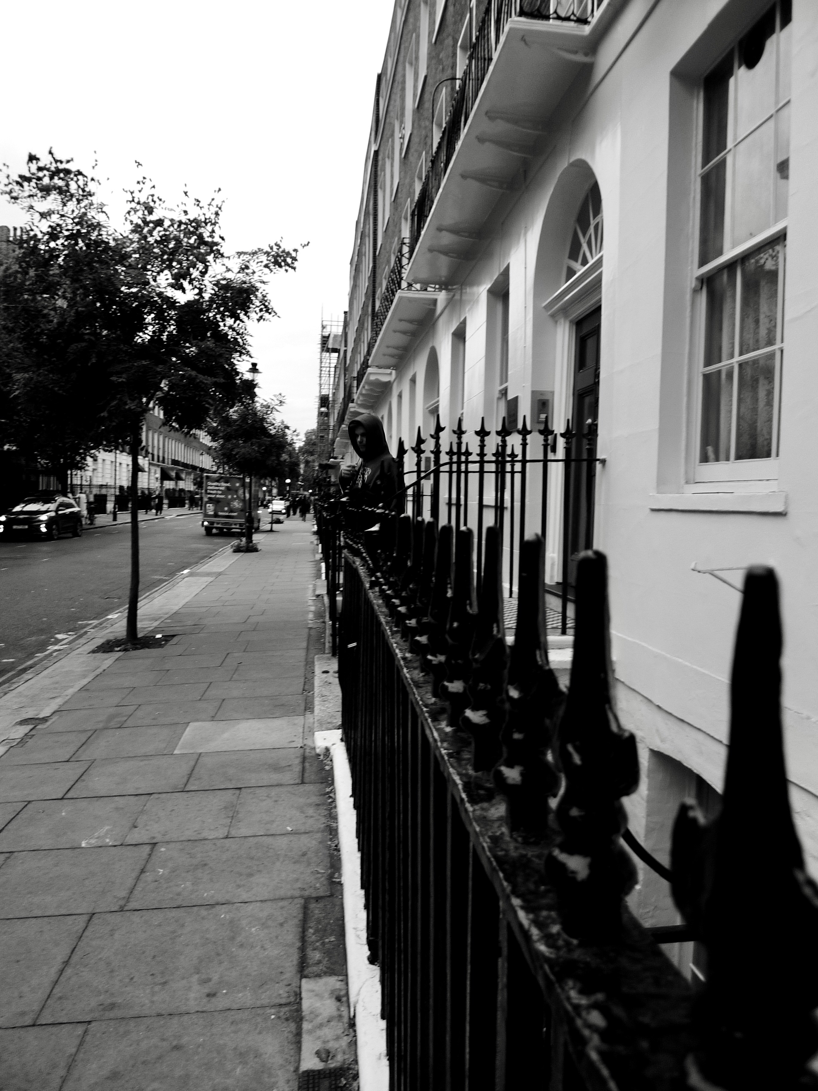 Monochrome street photograph of a lone figure leaning along a wrought-iron fence beside a row of townhouses. The scene emphasizes perspective and urban quiet.