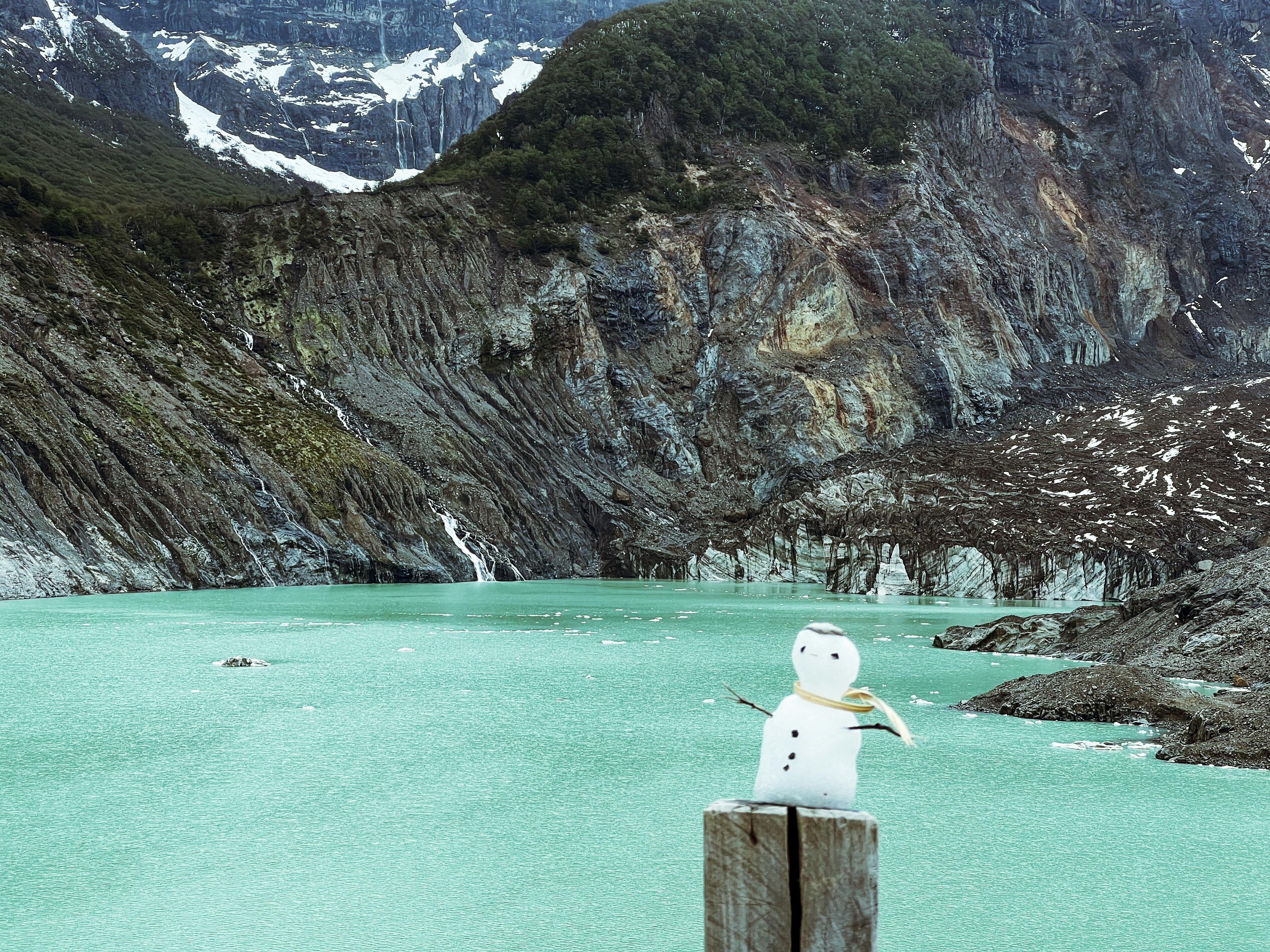 A snowman sitting on a post in front of a mountain lake photo – Free ...