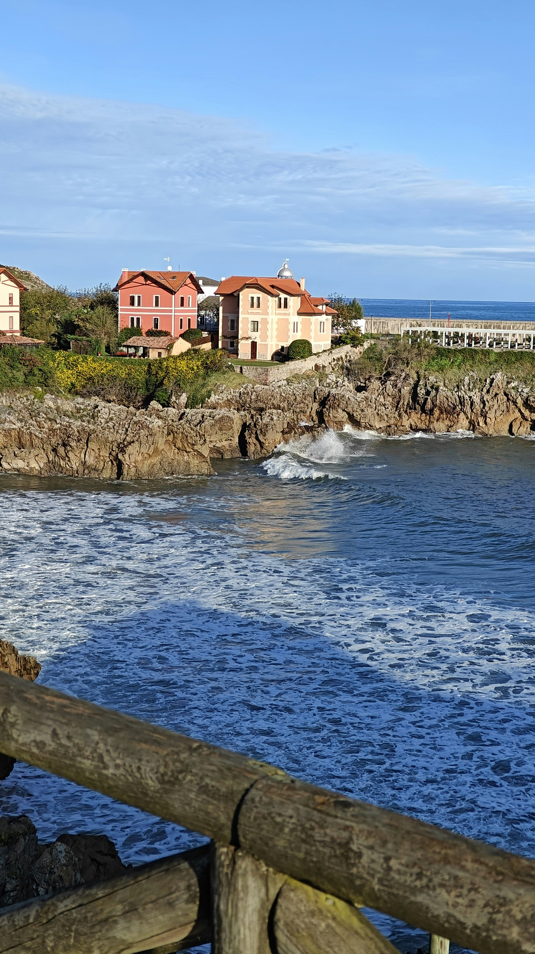Cliffside cottages with terracotta roofs overlook a rocky shoreline and blue sea, framed by a weathered wooden railing in the foreground.