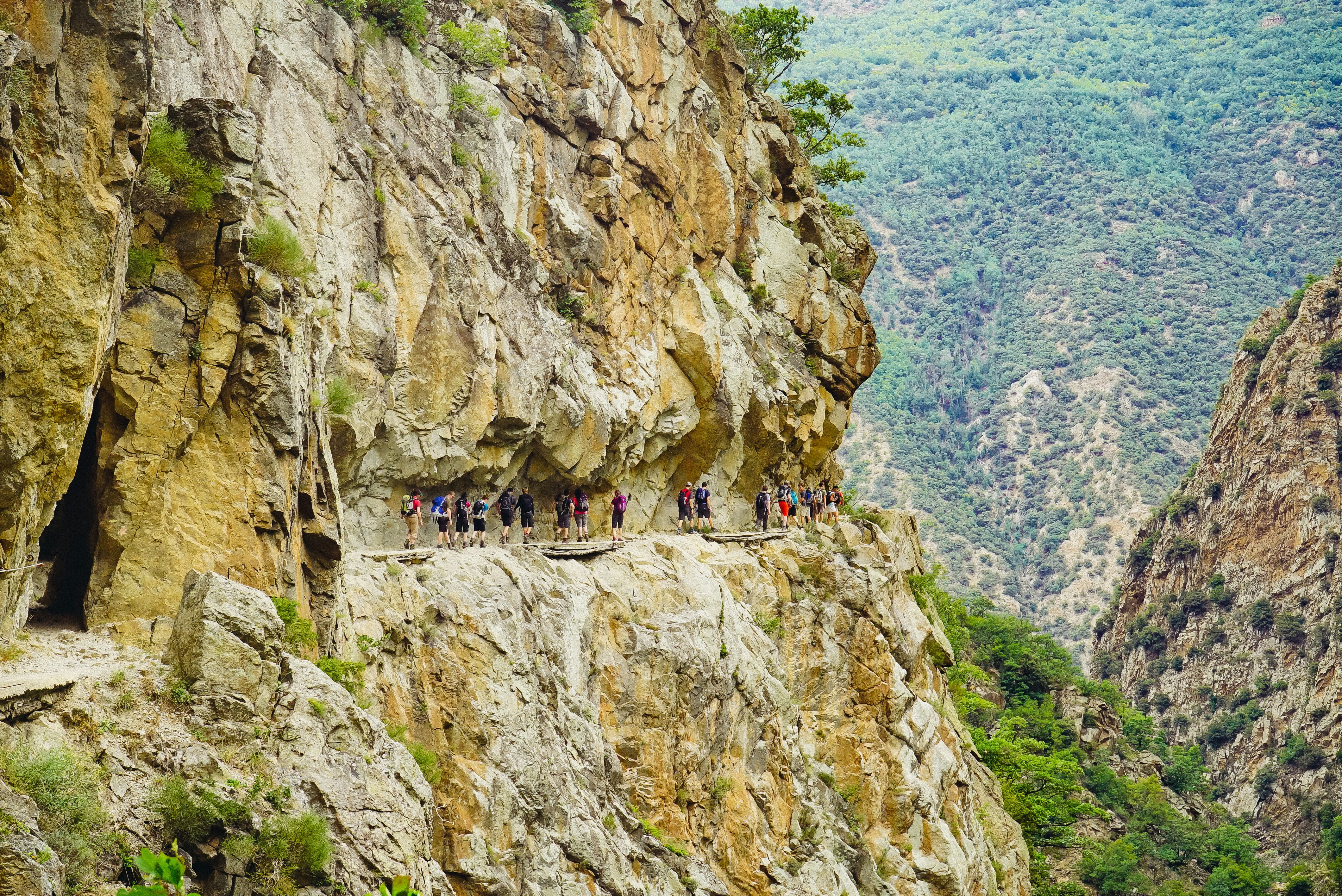 Hikers traverse a narrow trail carved into a rugged cliffside with lush green valleys below.