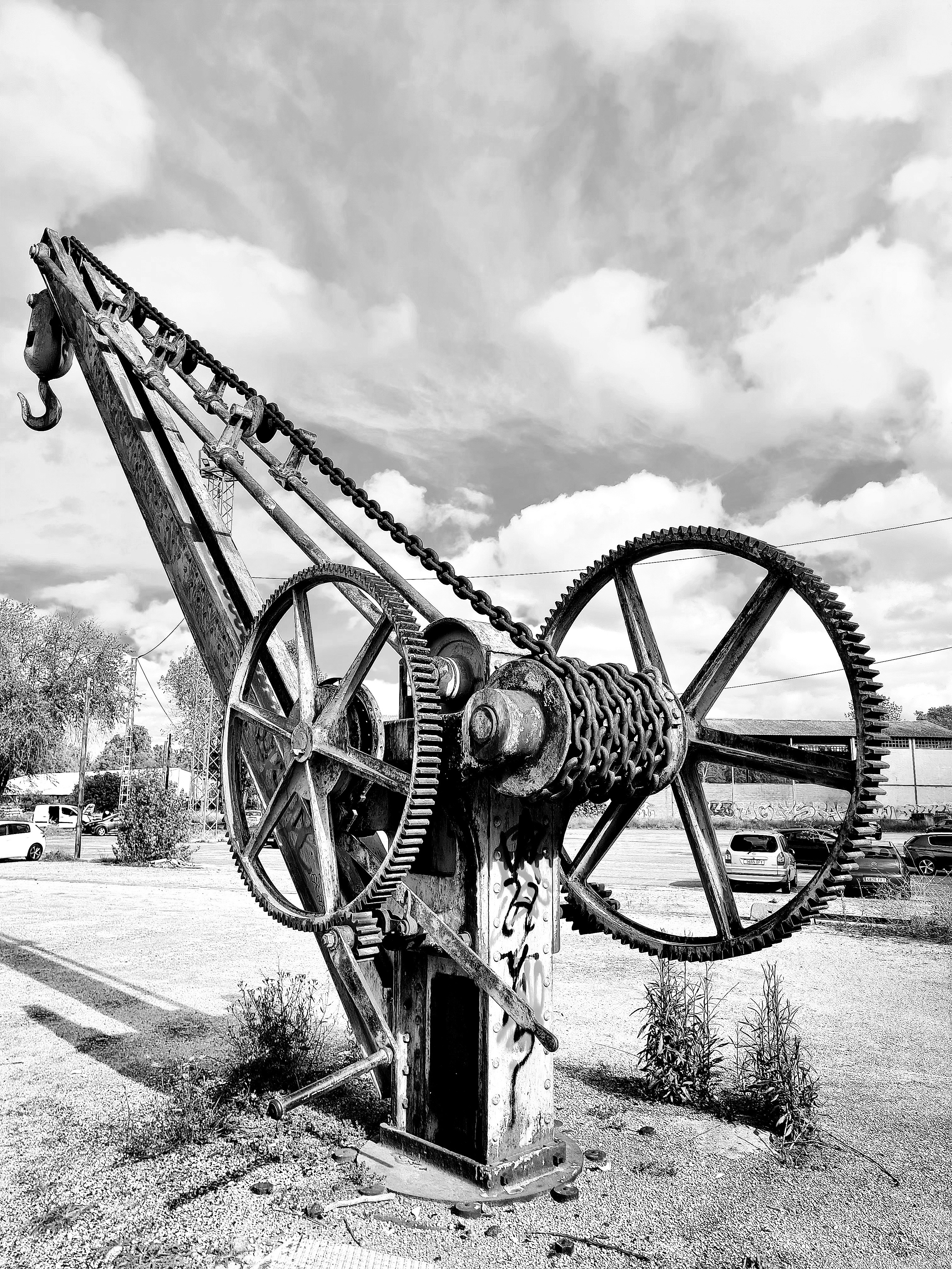 A weathered gear sculpture with large wheels and a chain dominates a parking lot. The black-and-white photograph emphasizes texture and industrial contrast against a cloudy sky.
