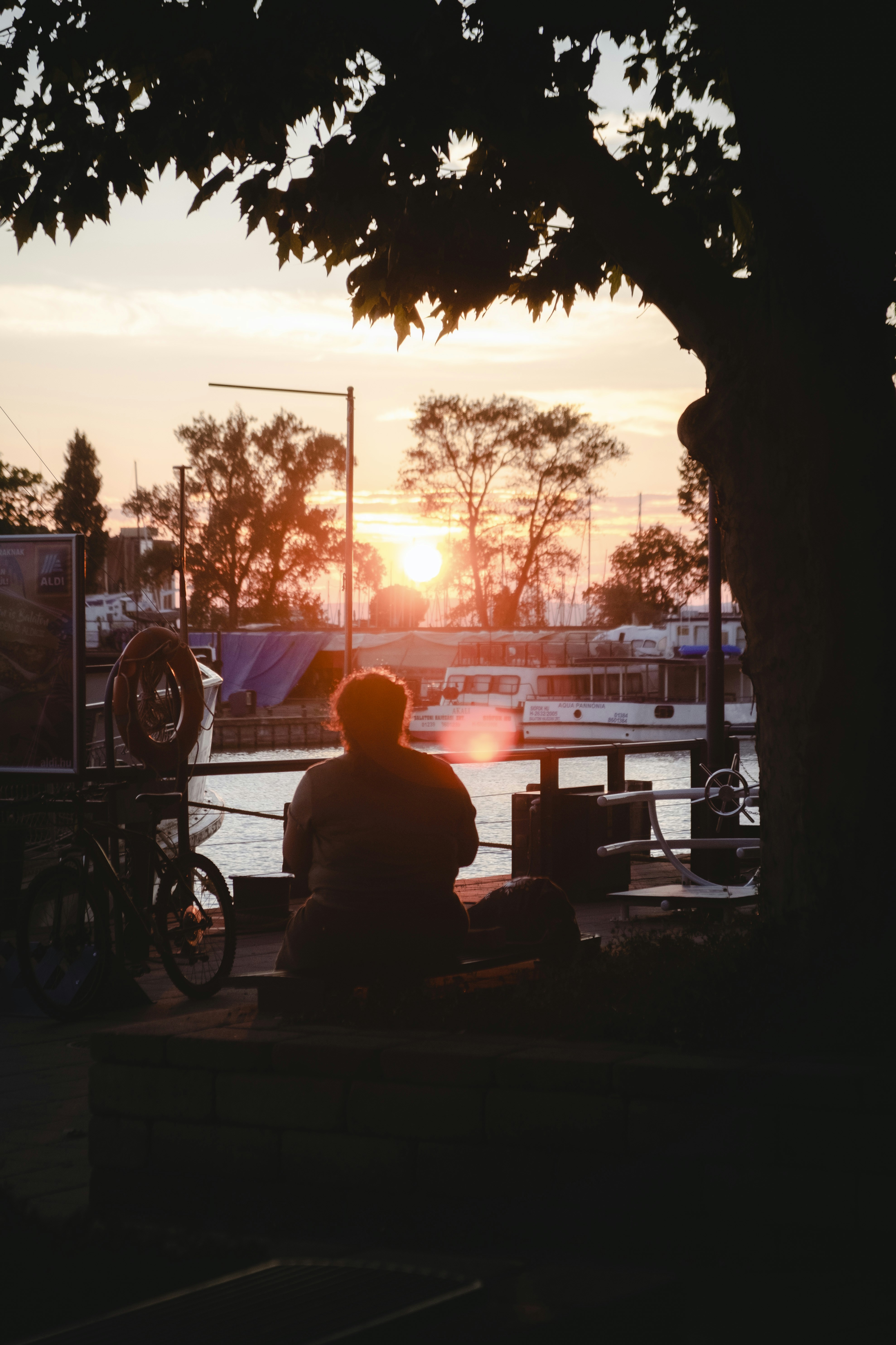 Silhouette of a person sitting by the waterfront, gazing at a vibrant sunset with boats in the background.