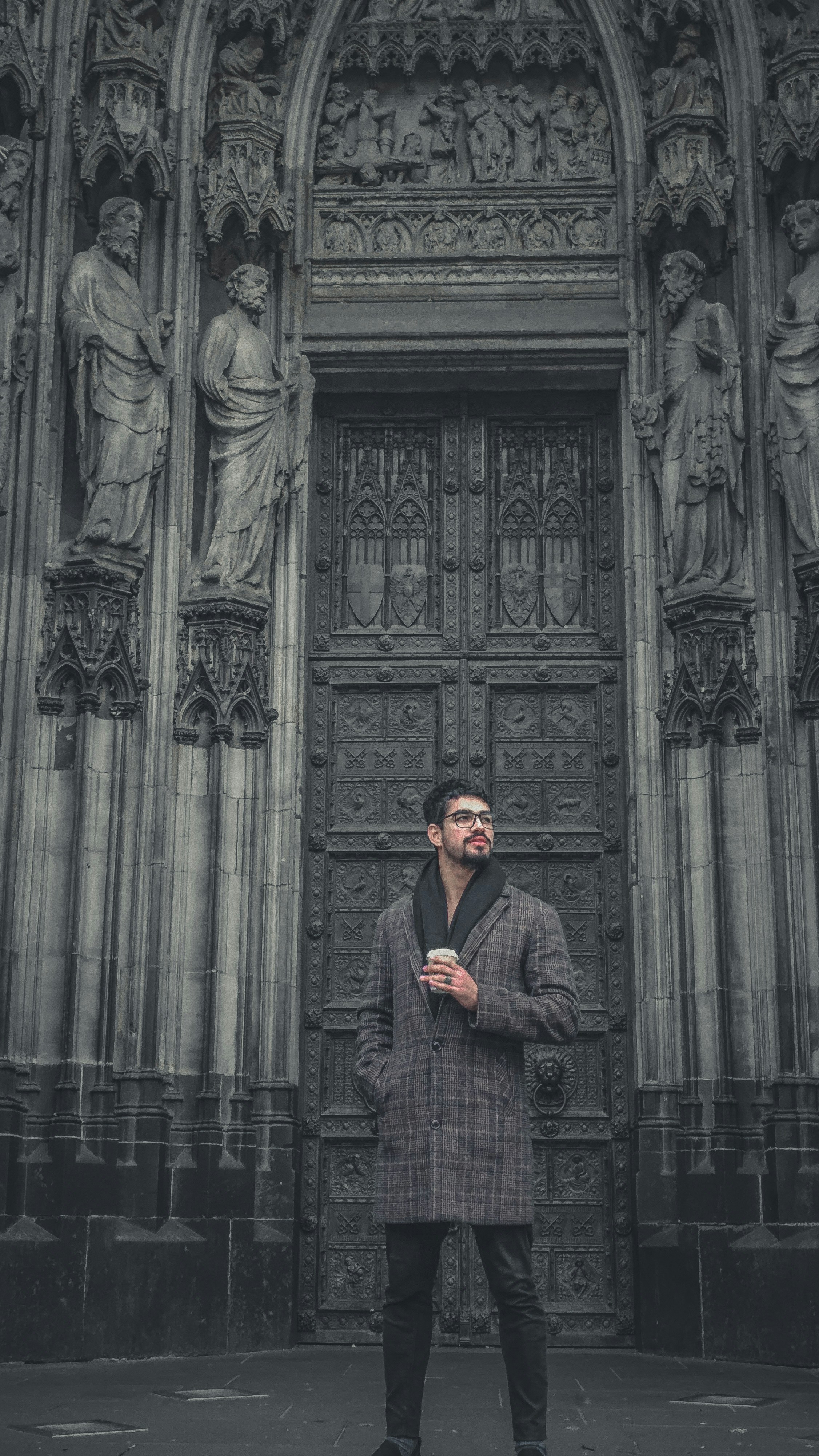 Photograph of a man in a plaid overcoat standing before an ornate cathedral doorway. The frame highlights the Gothic statues and carved details surrounding the entrance.