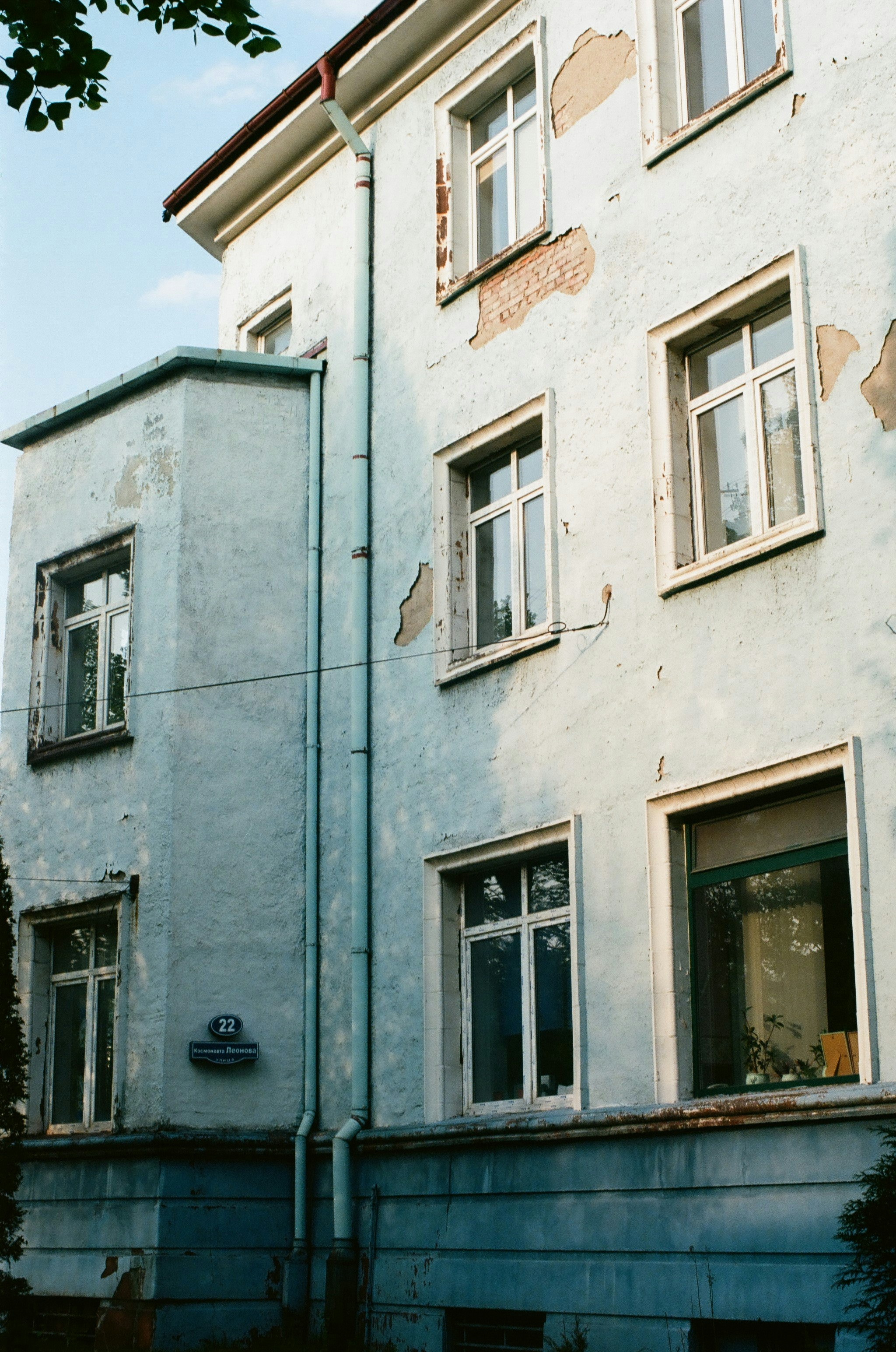 Weathered blue building facade with peeling plaster and exposed pipes. White-framed windows line the wall, catching the daylight.