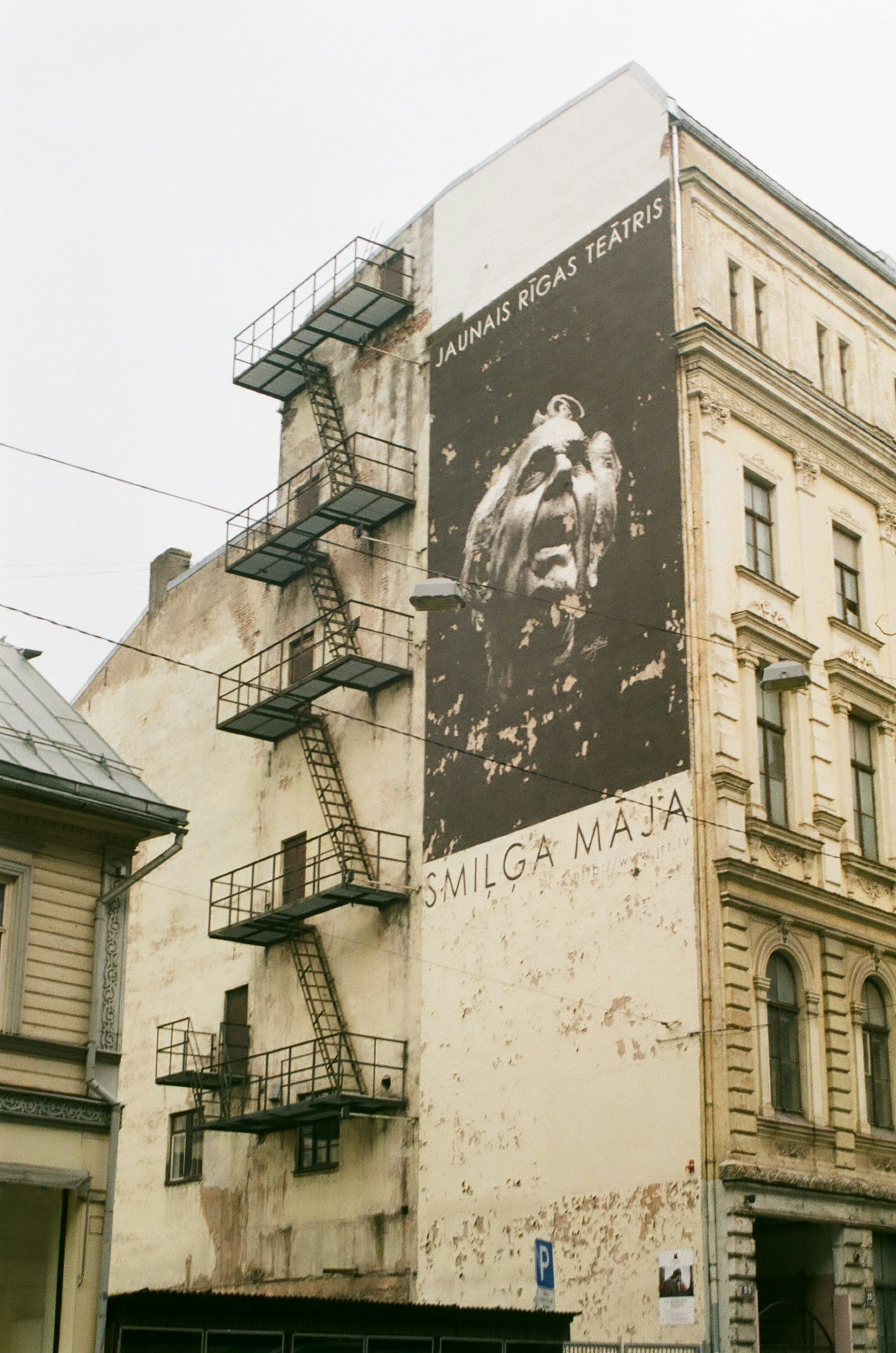 Photograph of a weathered urban facade featuring a large monochrome theater poster on the wall, with a rusted fire escape zigzagging downward.