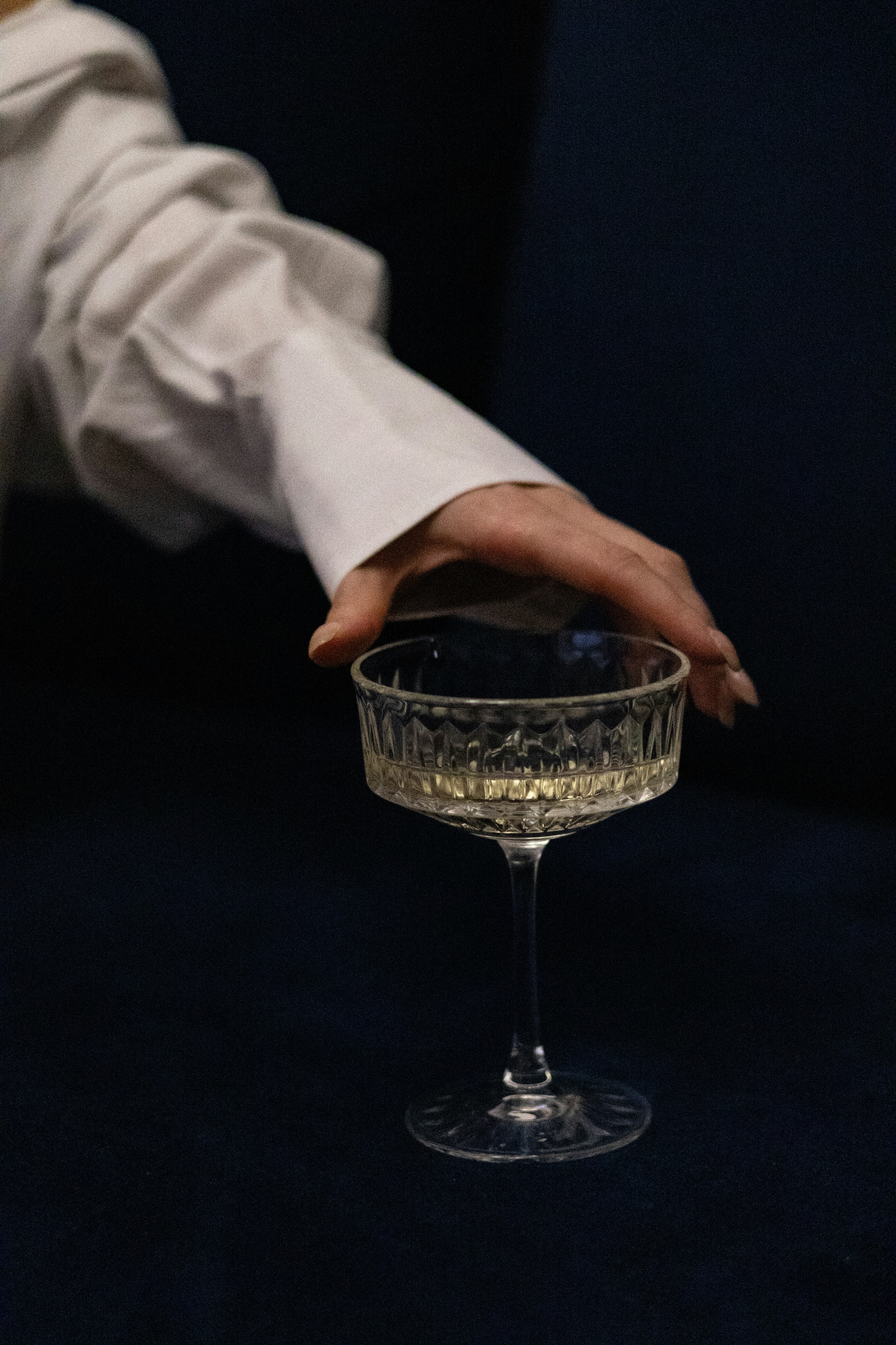 Hand hovers above a crystal coupe on a dark velvet surface, captured in moody low light.
