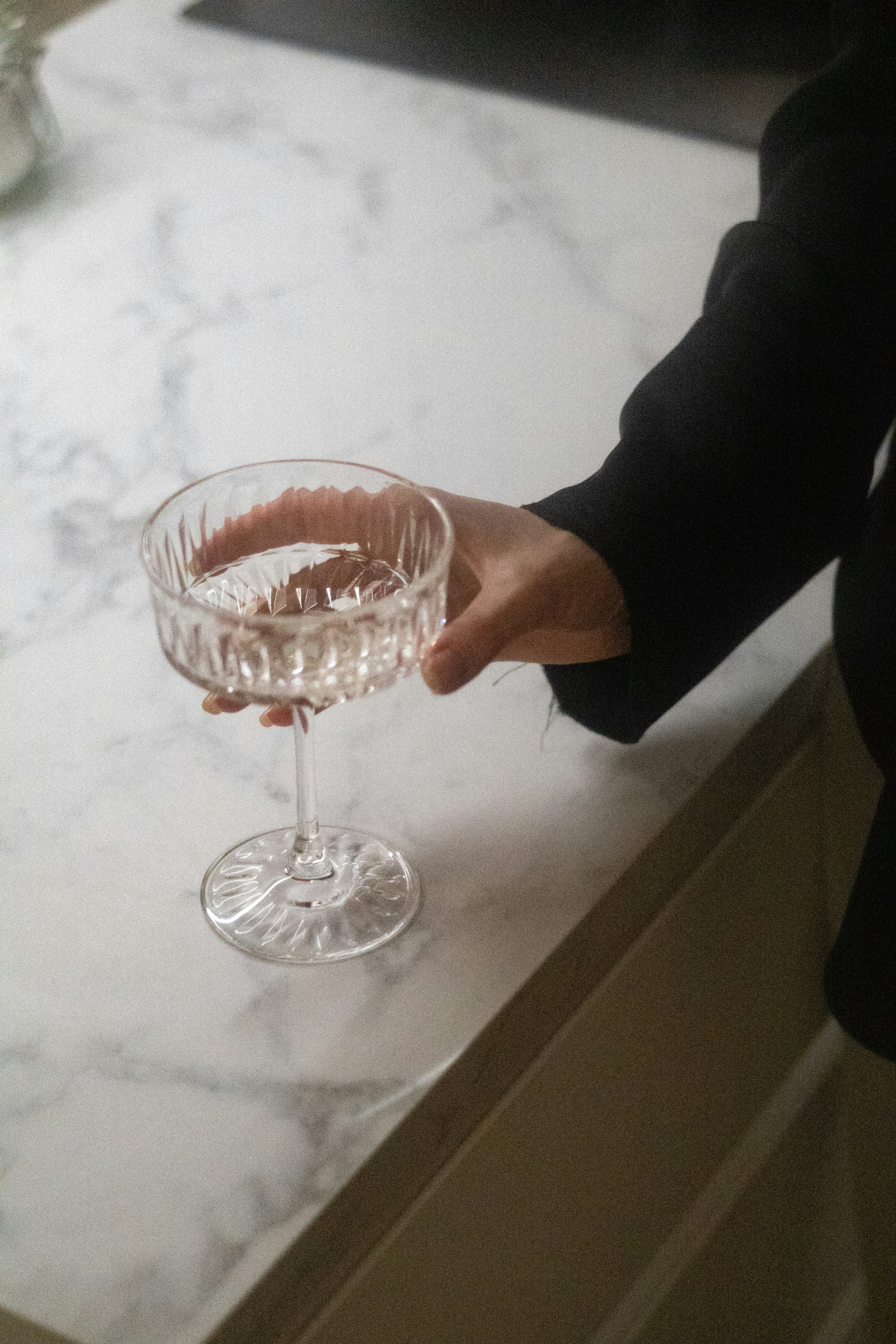 Photograph of a hand in a dark sleeve reaching toward a cut-crystal coupe glass on a white marble counter.