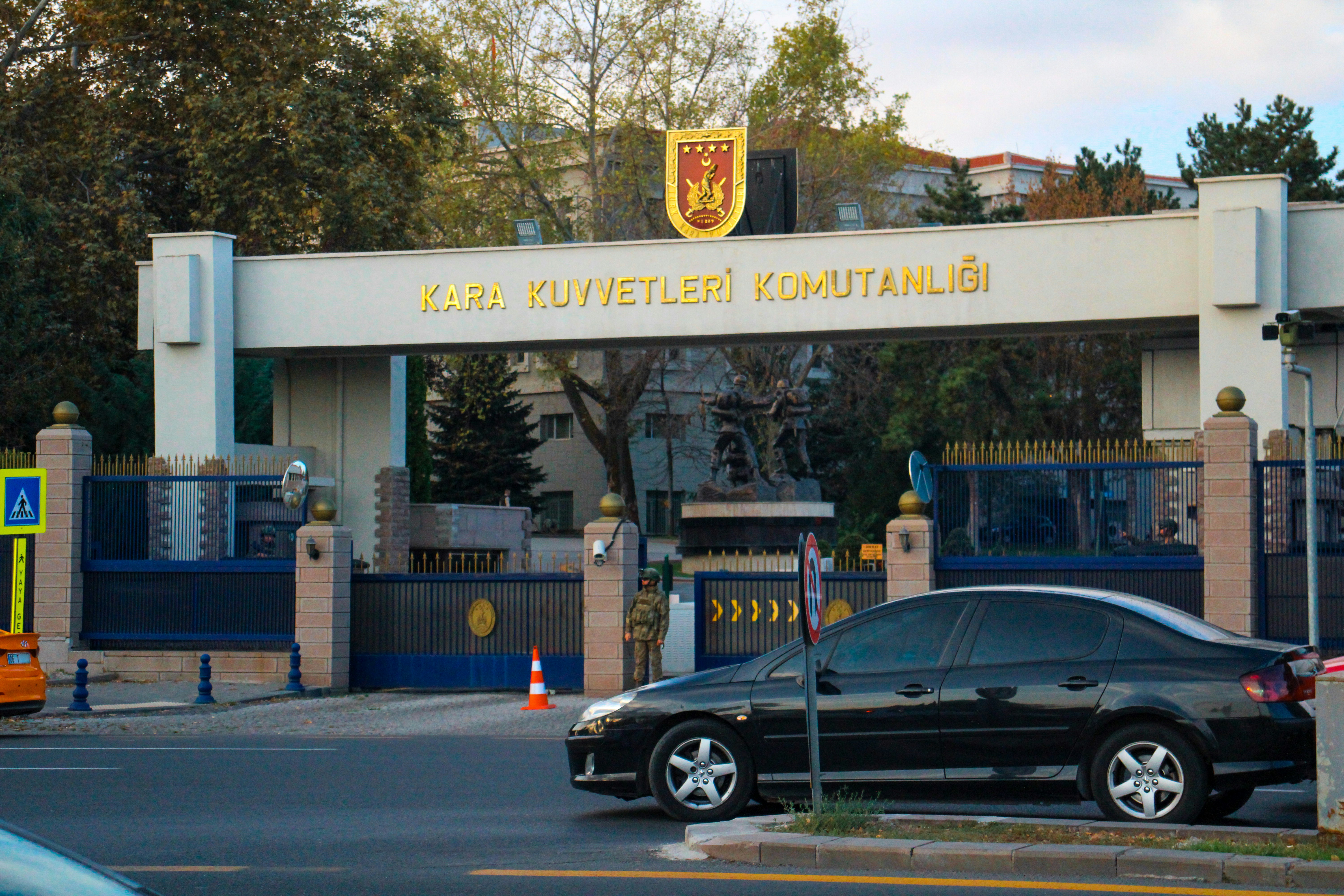 Dark sedan parked in front of a gated military compound with emblem above.