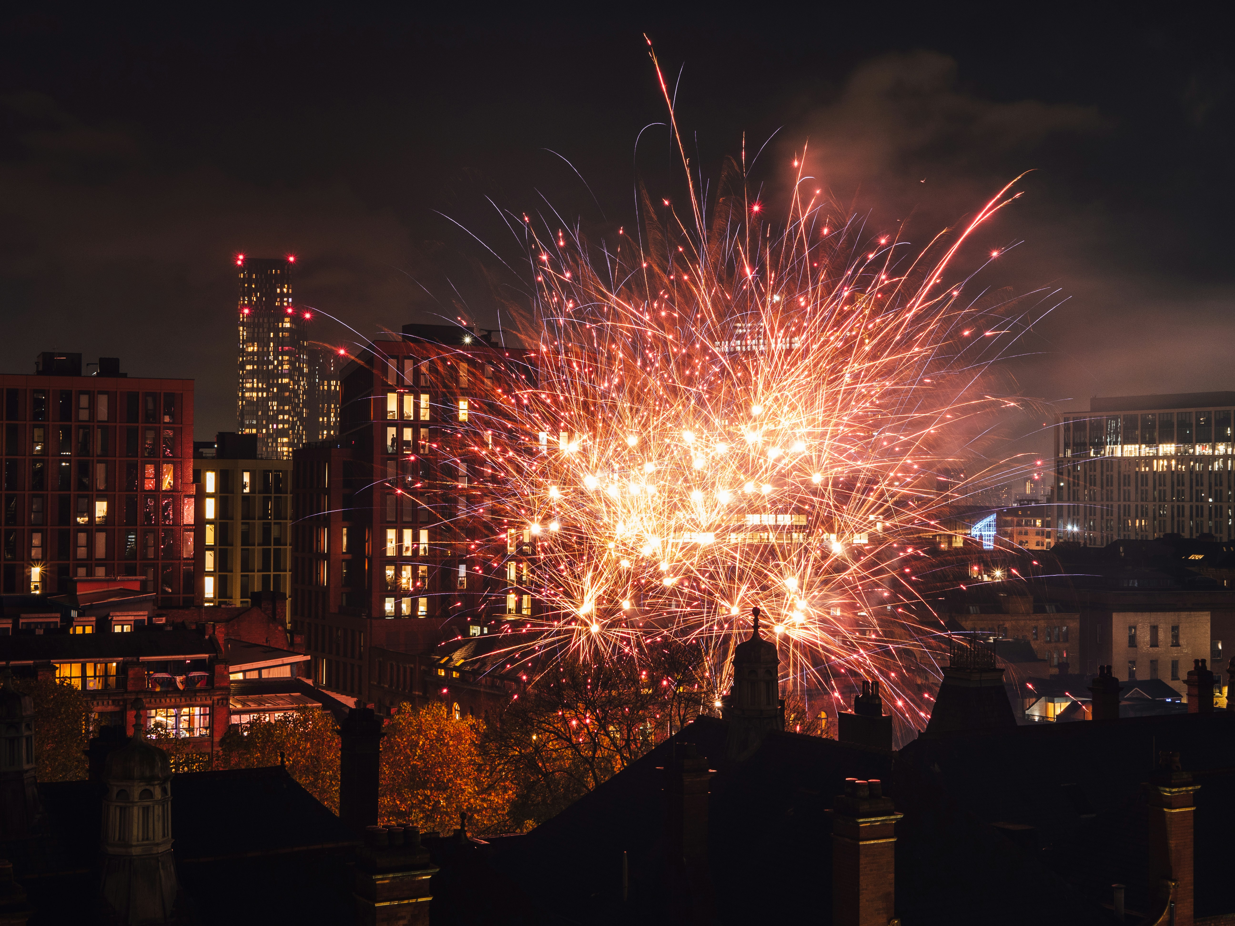 Fireworks illuminate city skyline at night, with glowing sparks and silhouetted buildings.