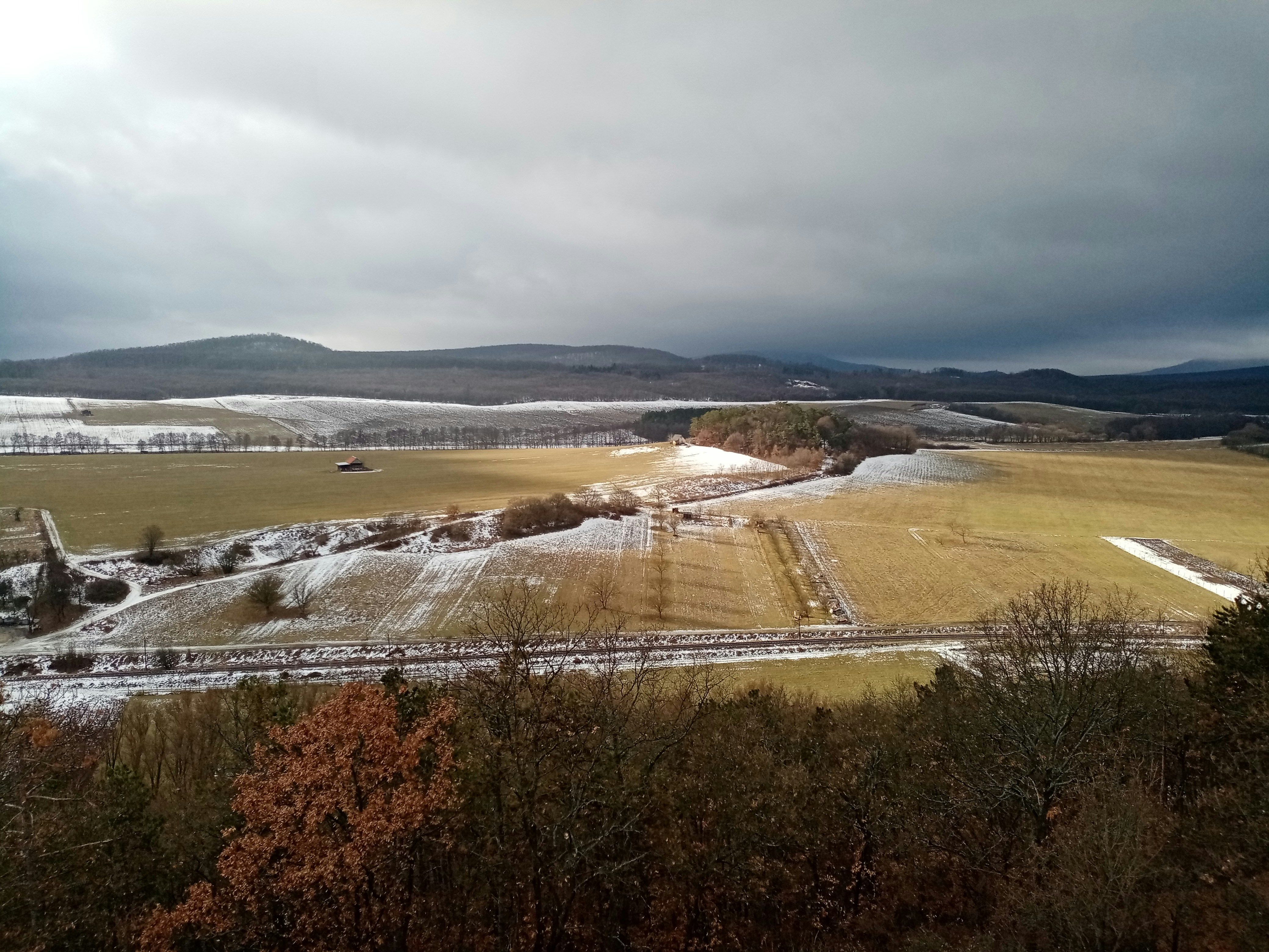 A winter landscape photograph showing snow patches across fields, a winding path, and distant hills beneath a cloudy sky.