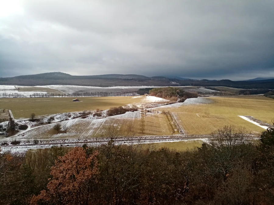 Wide rural landscape with patchy snow across fields, a winding road, and low hills in the distance beneath a dark cloudy sky. Trees frame the foreground.